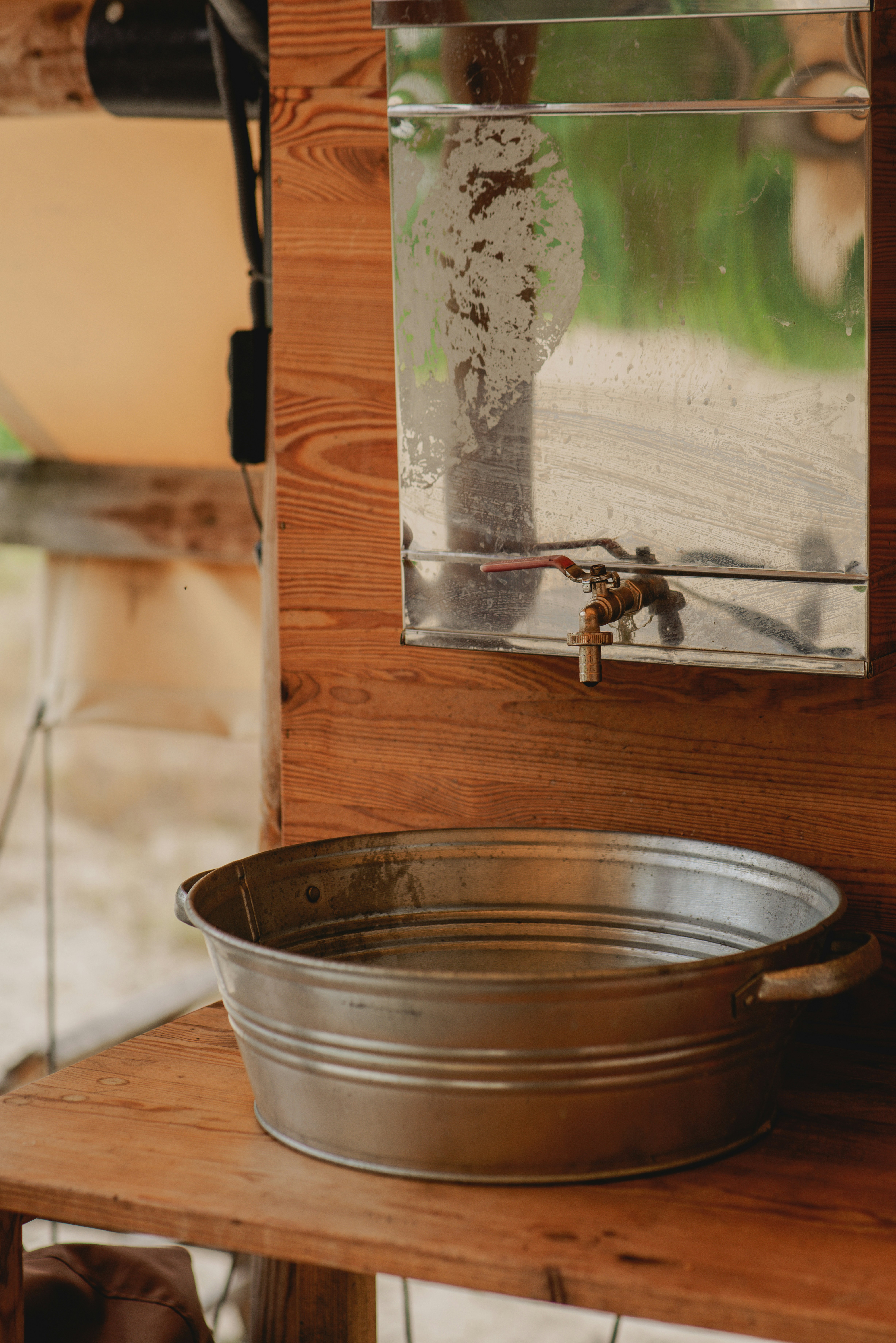 Metal wash basin positioned beneath a vintage water spout, reflecting the natural surroundings in a rustic setting.