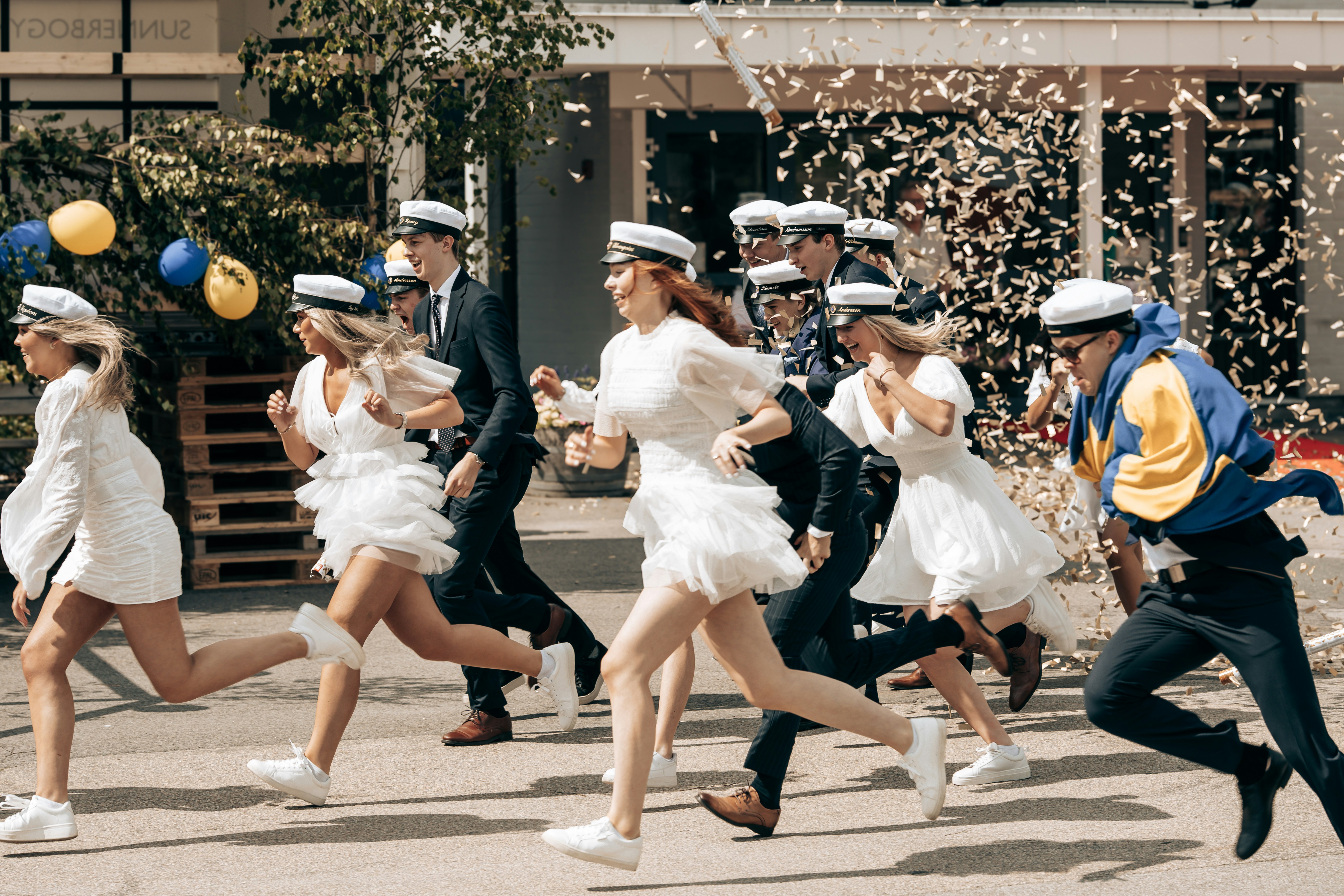 Group of young adults in white outfits and caps joyfully running together, surrounded by confetti and balloons during a festive event.