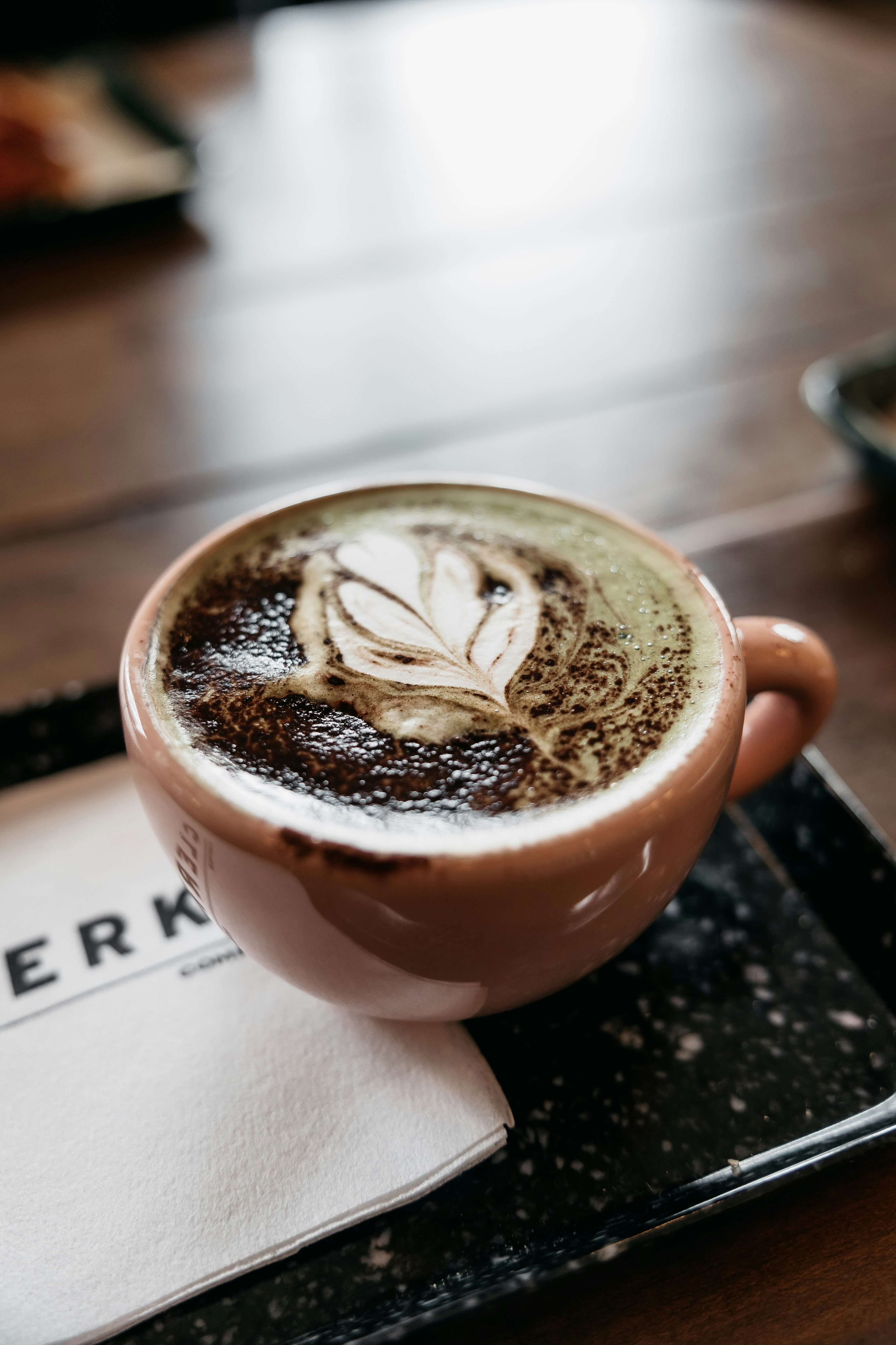 Matcha latte with intricate latte art atop, placed on a dark speckled tray beside a branded napkin.