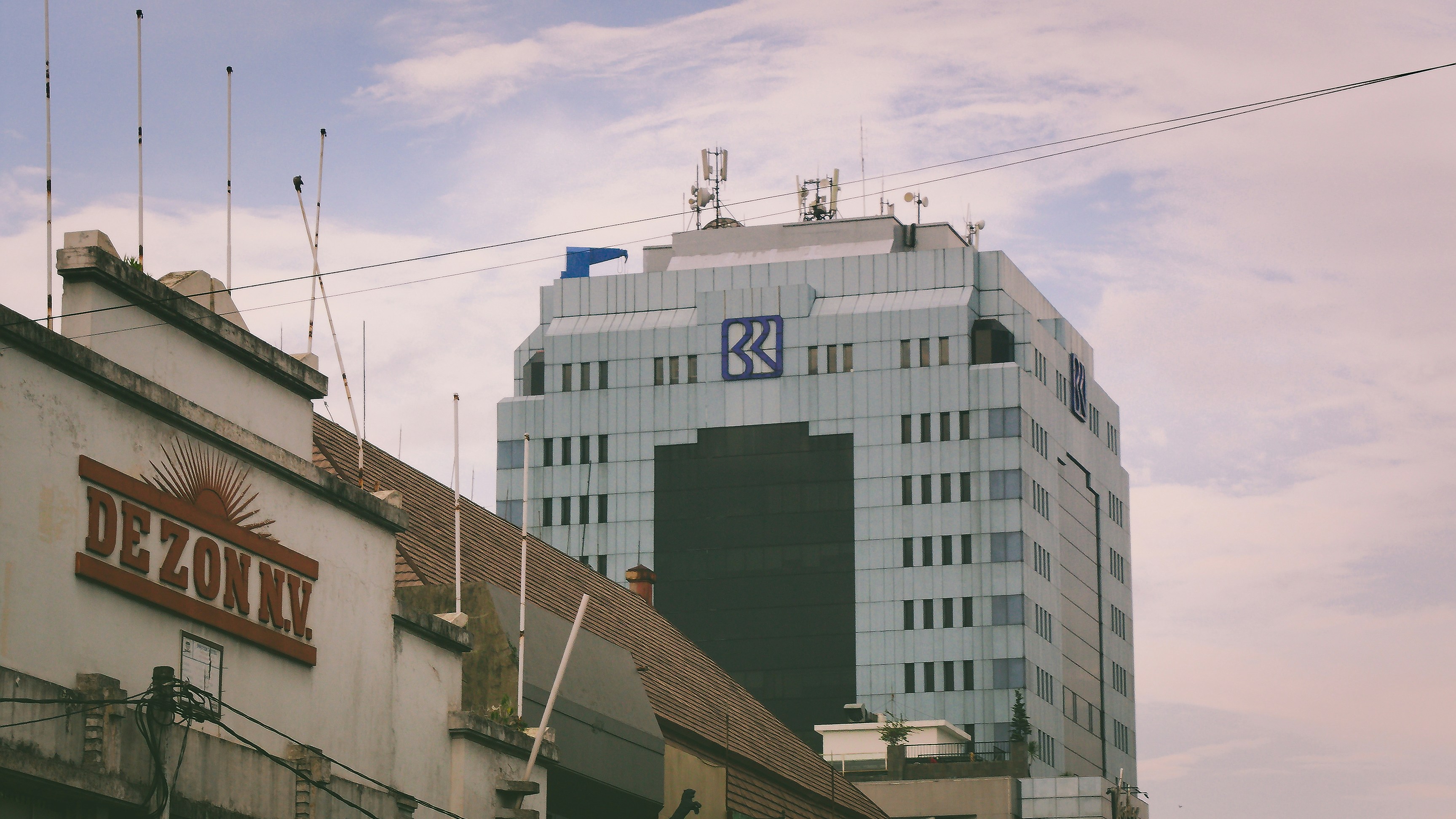 A view of the BRI (Bank Rakyat Indonesia) tower peeking behind old urban buildings with visible signs of age, including one marked “DEZON N.V.” The juxtaposition of modern and colonial architecture, power cables, and rooftop antennas gives this street scene a gritty yet nostalgic urban charm. | A modern building rises above older ones.
