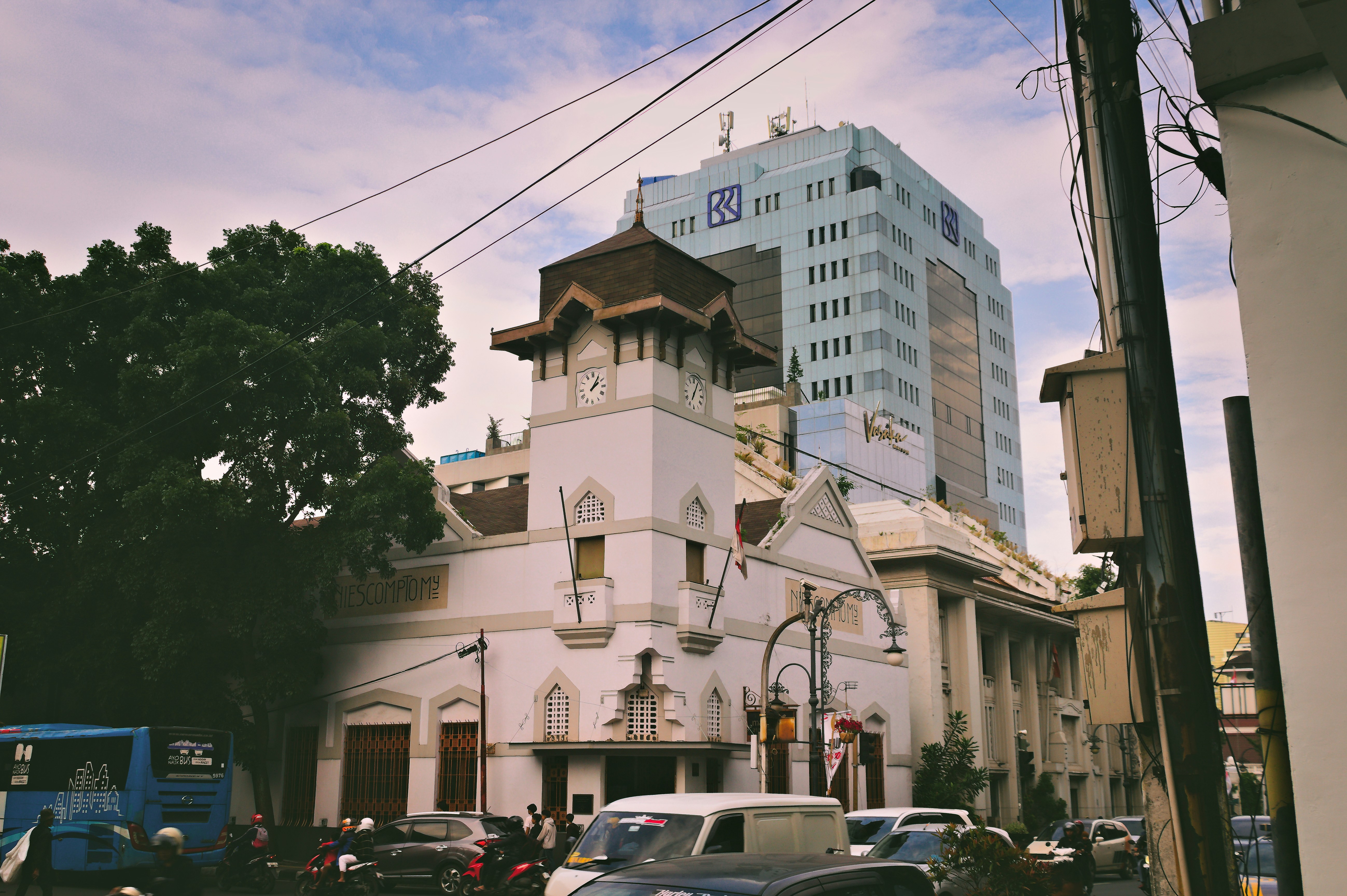 A classic street scene blending colonial architecture and modern urban life. In the foreground stands the Eks Nederlandsch Indische Escompto Maatschappij building, a historical Dutch-era structure with a central clock tower, white walls, and arched windows. Behind it rises a modern skyscraper marked with the "BRI" logo, representing Bank Rakyat Indonesia. The contrast between old and new is striking—colonial elegance meets corporate minimalism. The streets are lively with cars, motorcycles, pedestrians, and a blue city bus, reflecting the dynamic rhythm of Bandung’s city life. | Old building stands in front of a modern one.