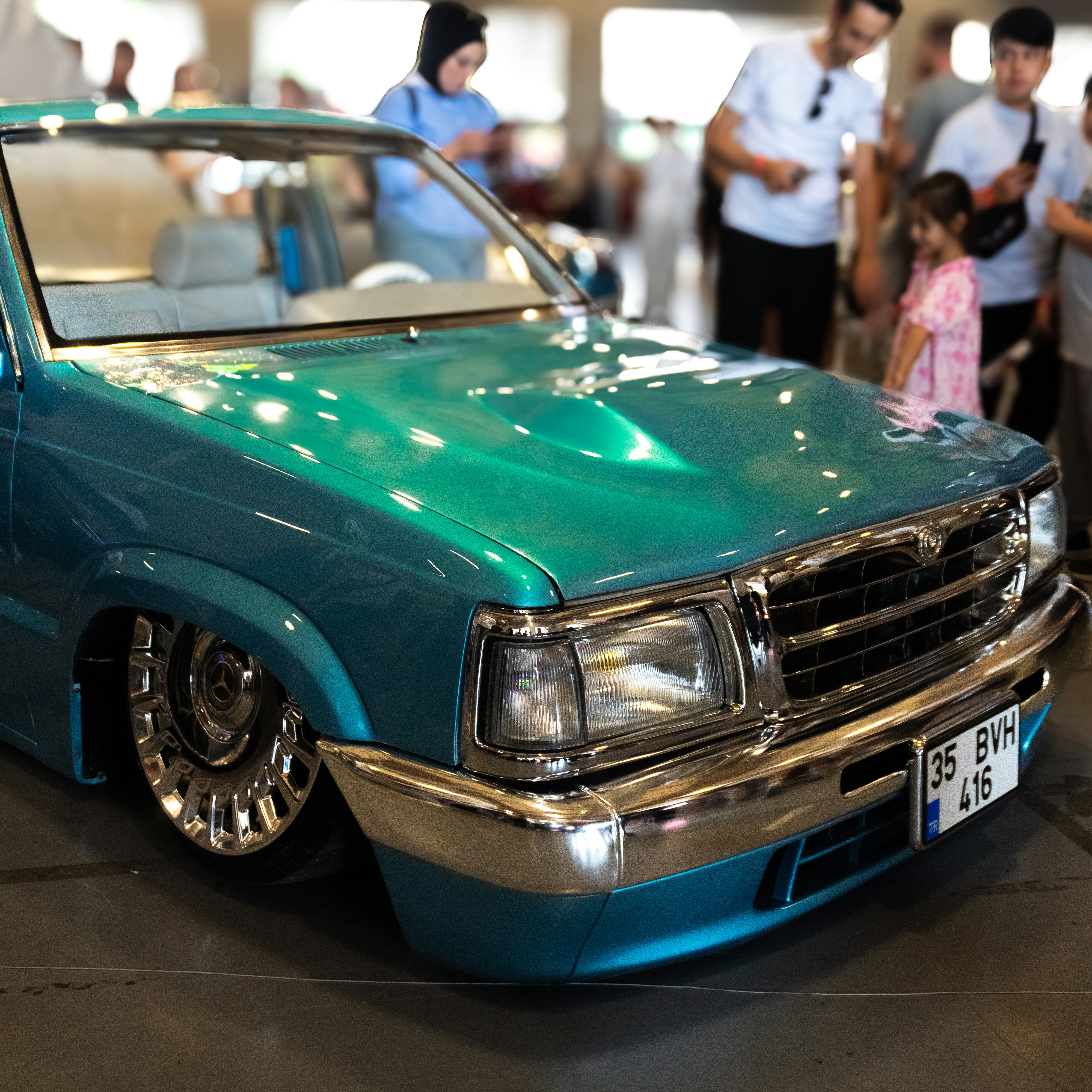 Custom turquoise car with a lowered stance and chrome wheels, surrounded by onlookers at an automotive event.