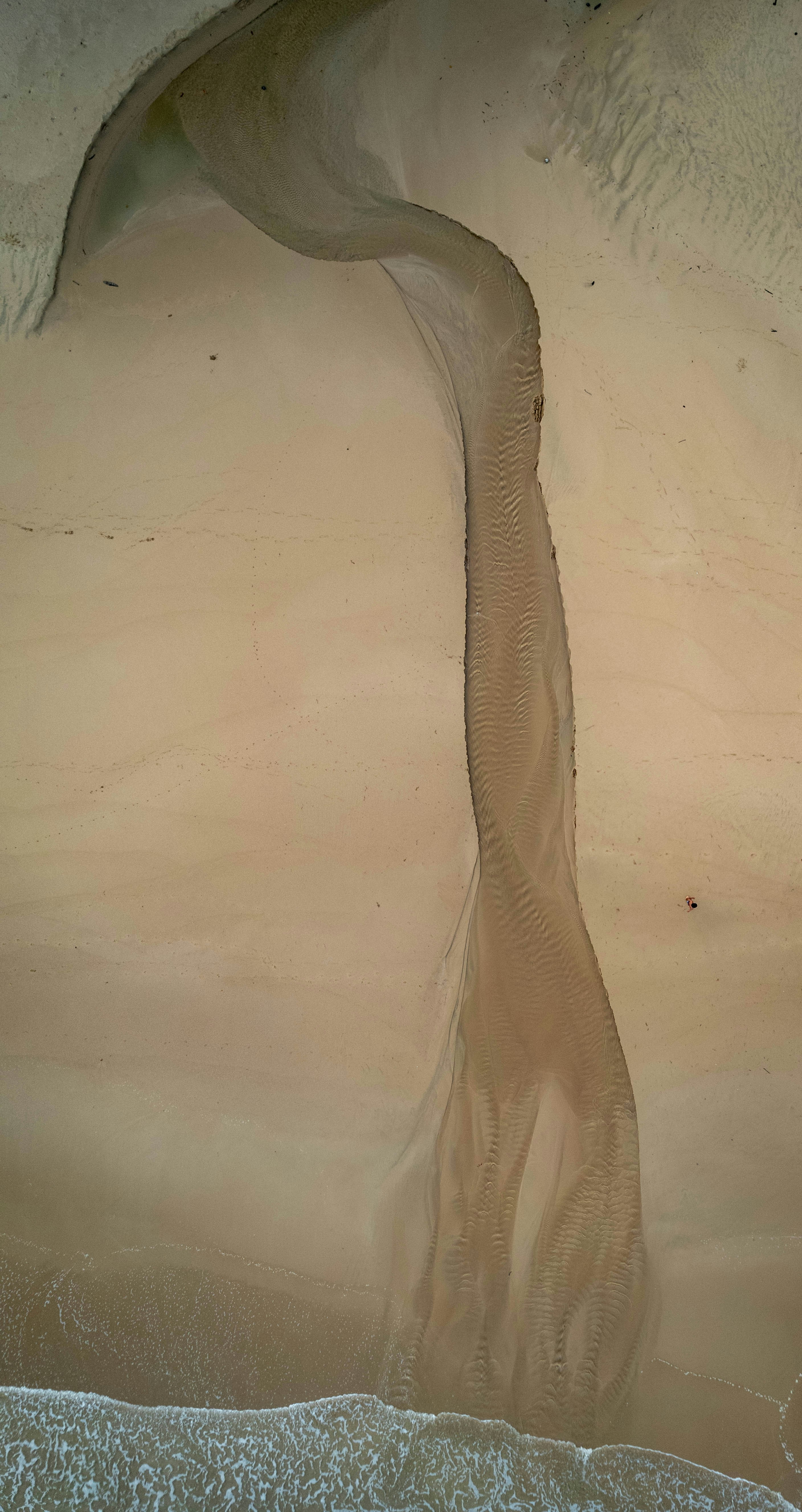 Aerial views of a tropical beach show waves breaking along the shoreline, bordered by dense green vegetation and sandy stretches. In some images, small estuaries carve through the sand into the ocean. Surfers and surfboards are visible in the shallows, navigating the whitewater near the beach. | A stream flows to the ocean through the sand.