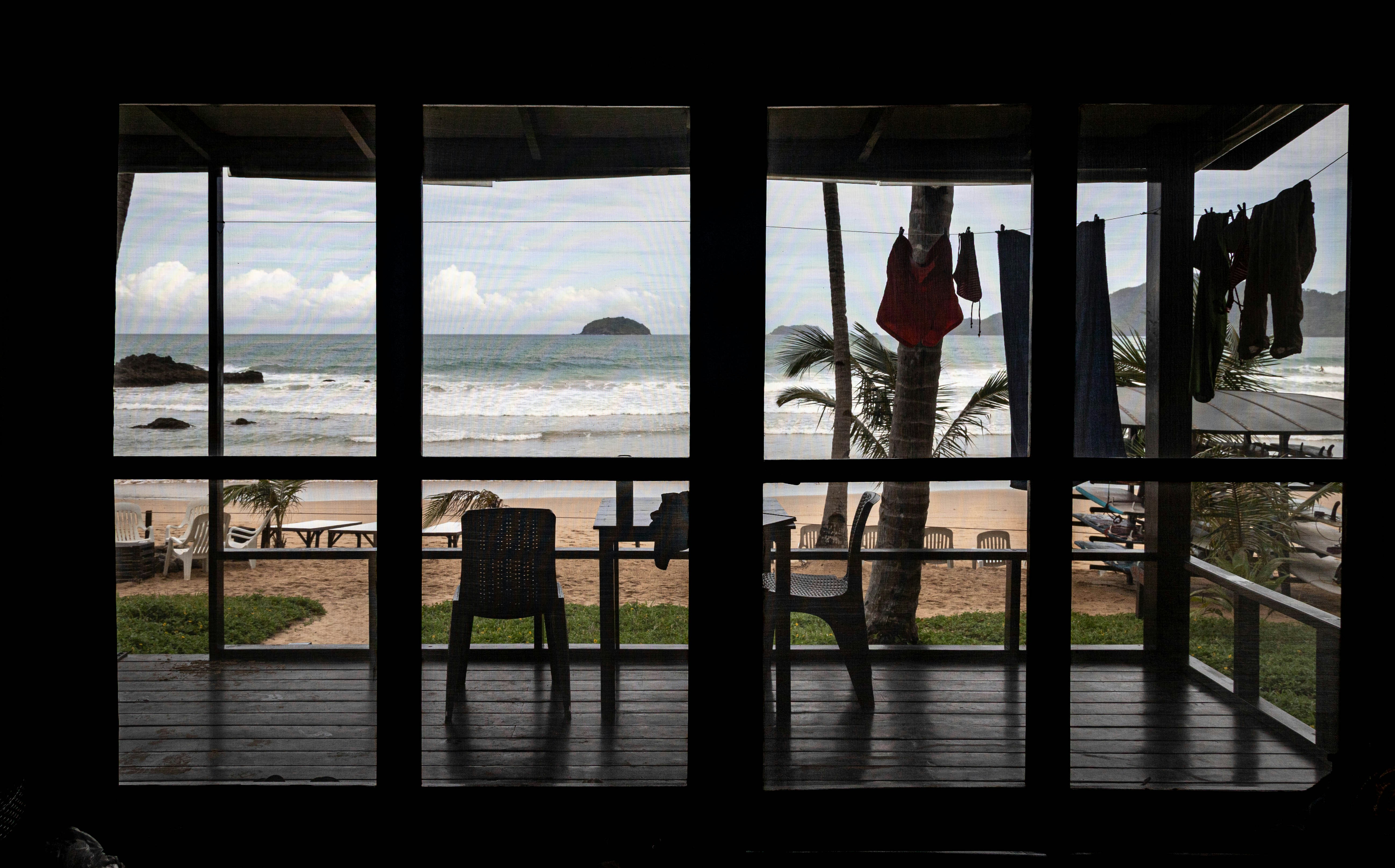 View of a beach scene from inside a cabin, featuring a chair and clothes hanging on a line, with ocean waves and a distant island visible.