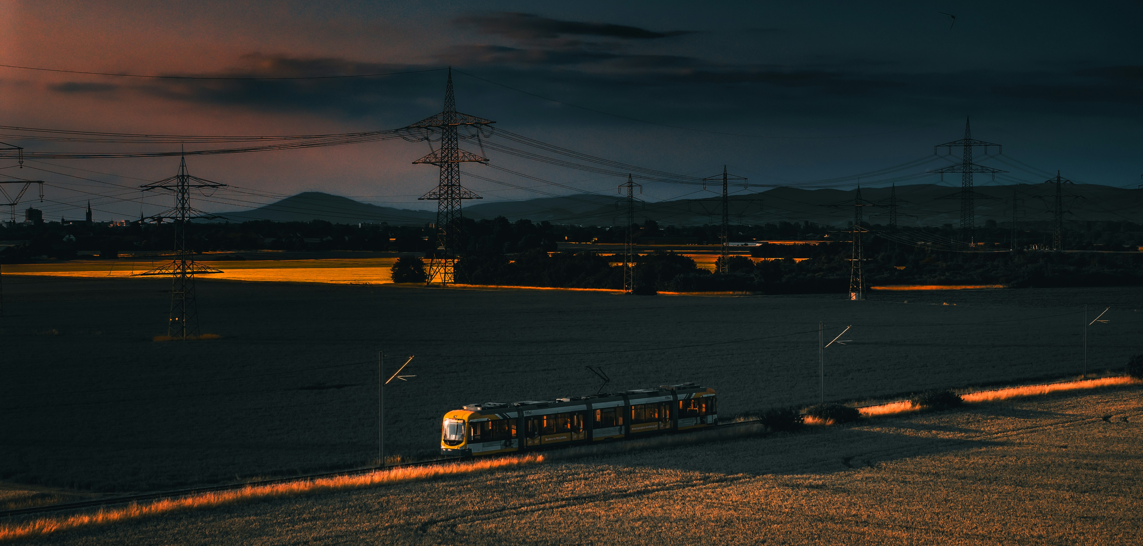 A tram glides along tracks in a vast golden field under a dramatic twilight sky, framed by distant mountains and power lines.