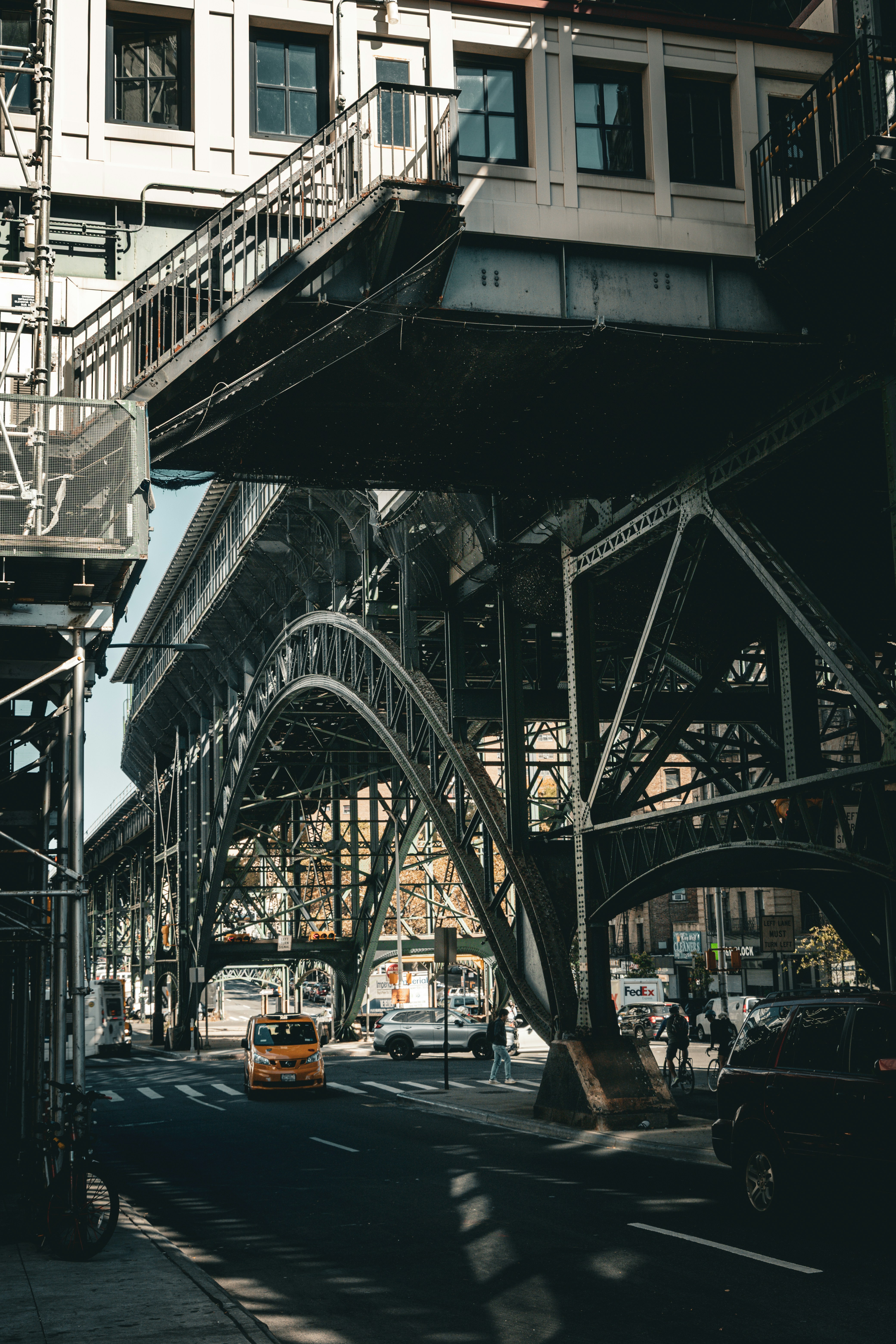 Intricate steel arches create a dynamic interplay of light and shadow in a bustling city scene, showcasing urban architecture. A yellow taxi navigates the street below.
