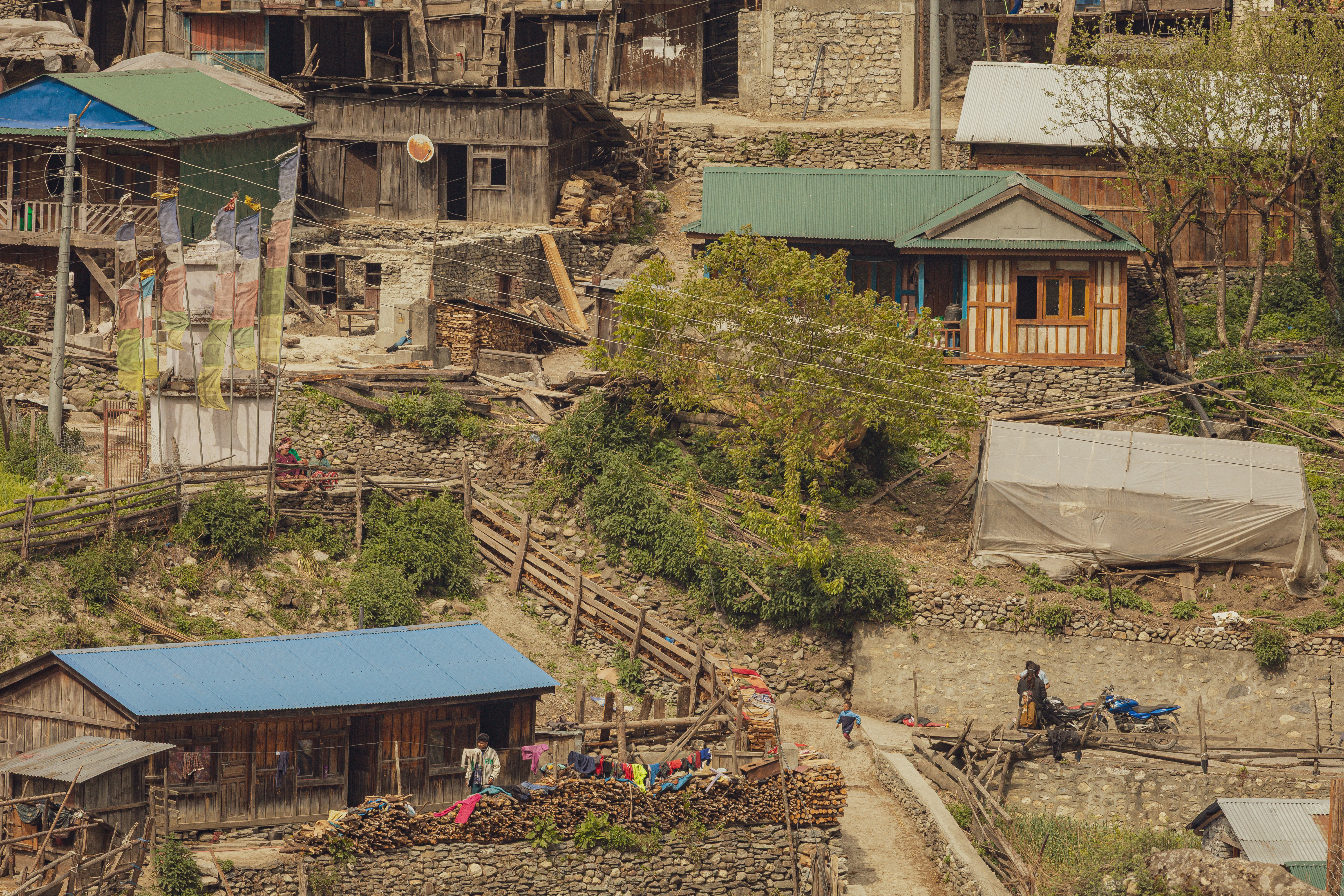 A serene mountain village showcasing rustic wooden houses, a vibrant blue roof, and a glimpse of daily life with villagers and livestock. The landscape reflects a blend of nature and human habitation.