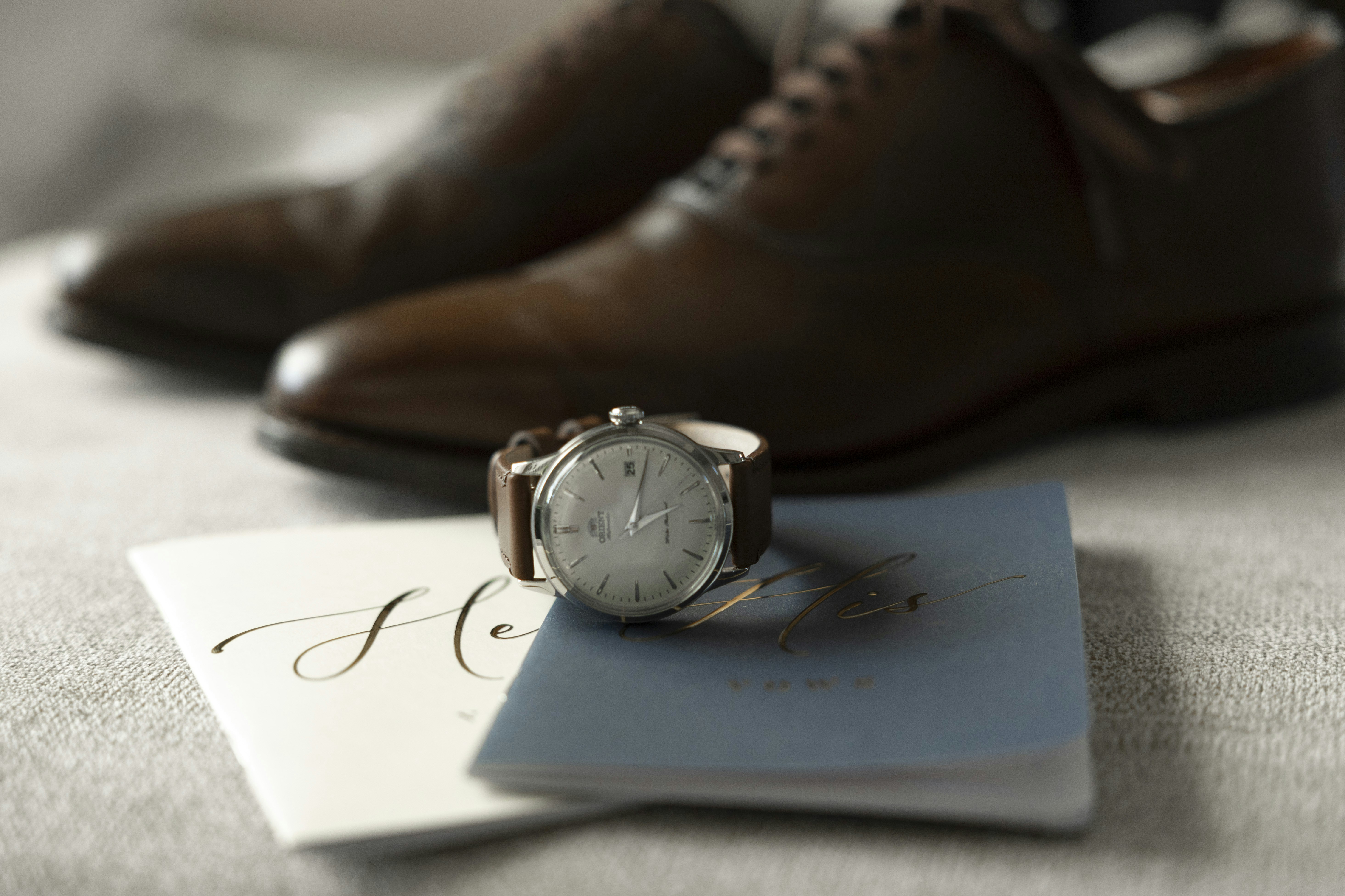 Classic wristwatch resting on a soft surface next to a pair of polished brown shoes and elegant stationery.