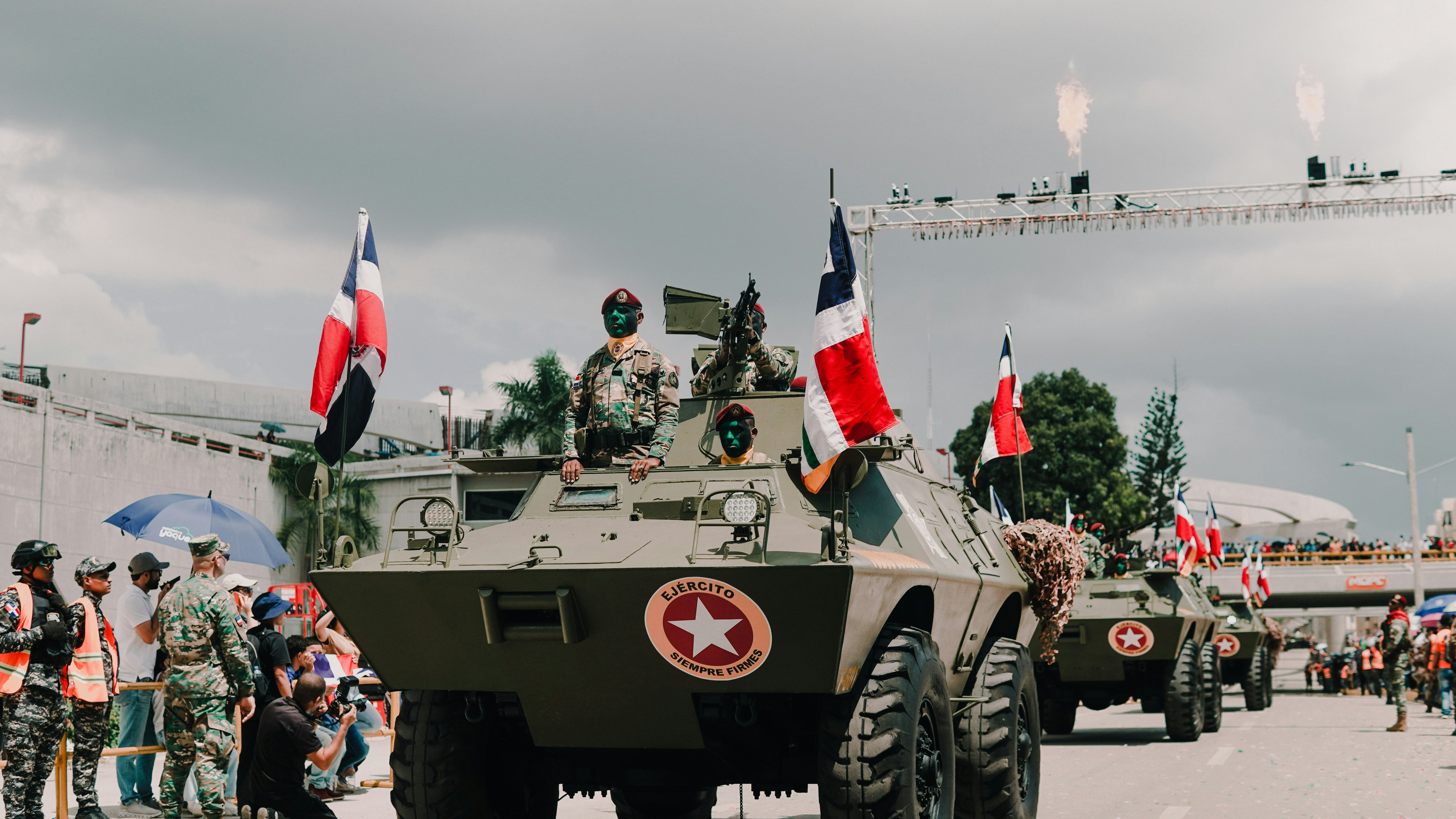 Military vehicles adorned with flags parade down a street, showcasing a blend of patriotism and tradition during a national celebration.
