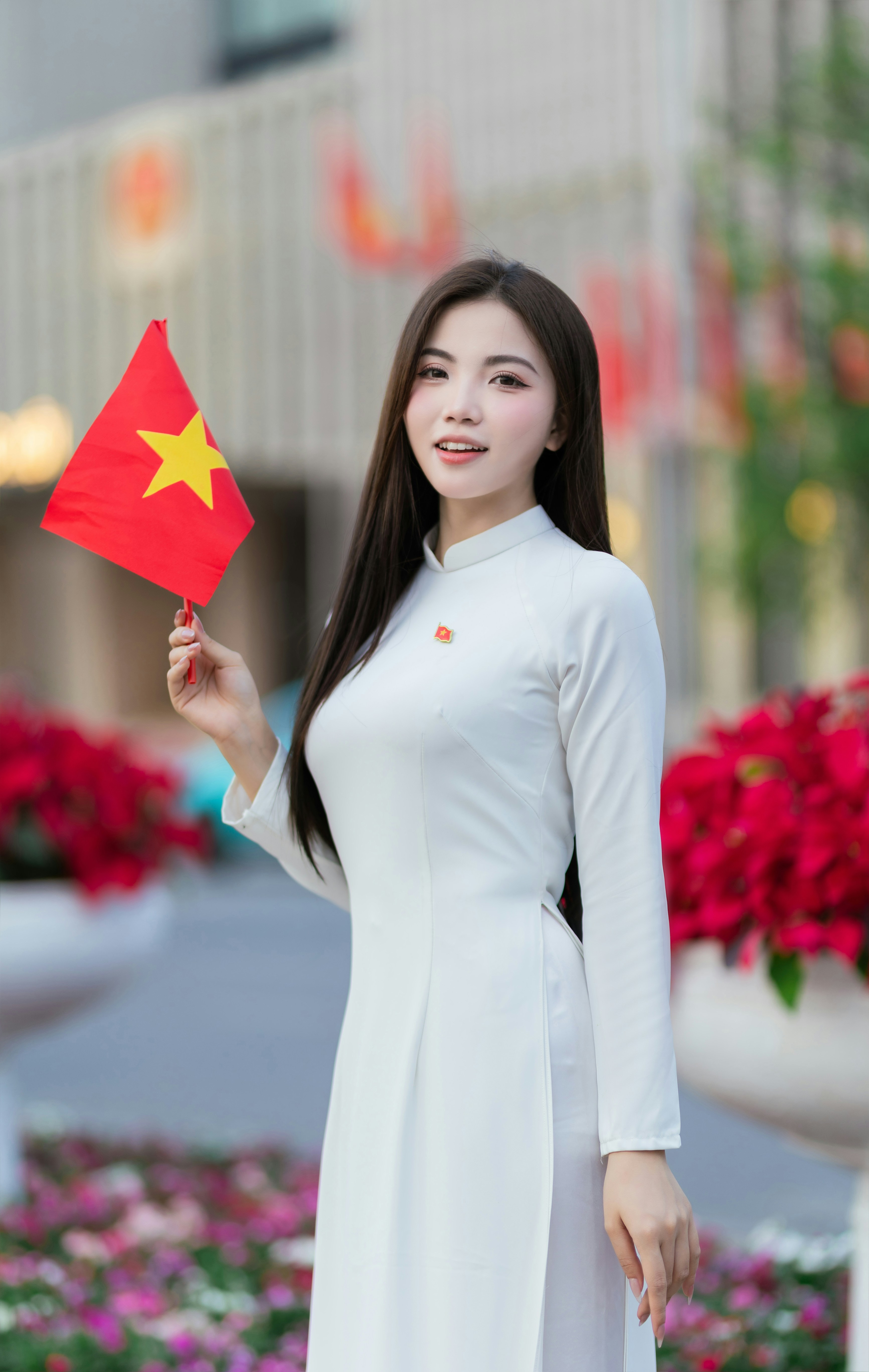 A young woman in a traditional white ao dai holds a red flag with a yellow star, symbolizing Vietnamese heritage, amidst vibrant floral displays in an urban setting.