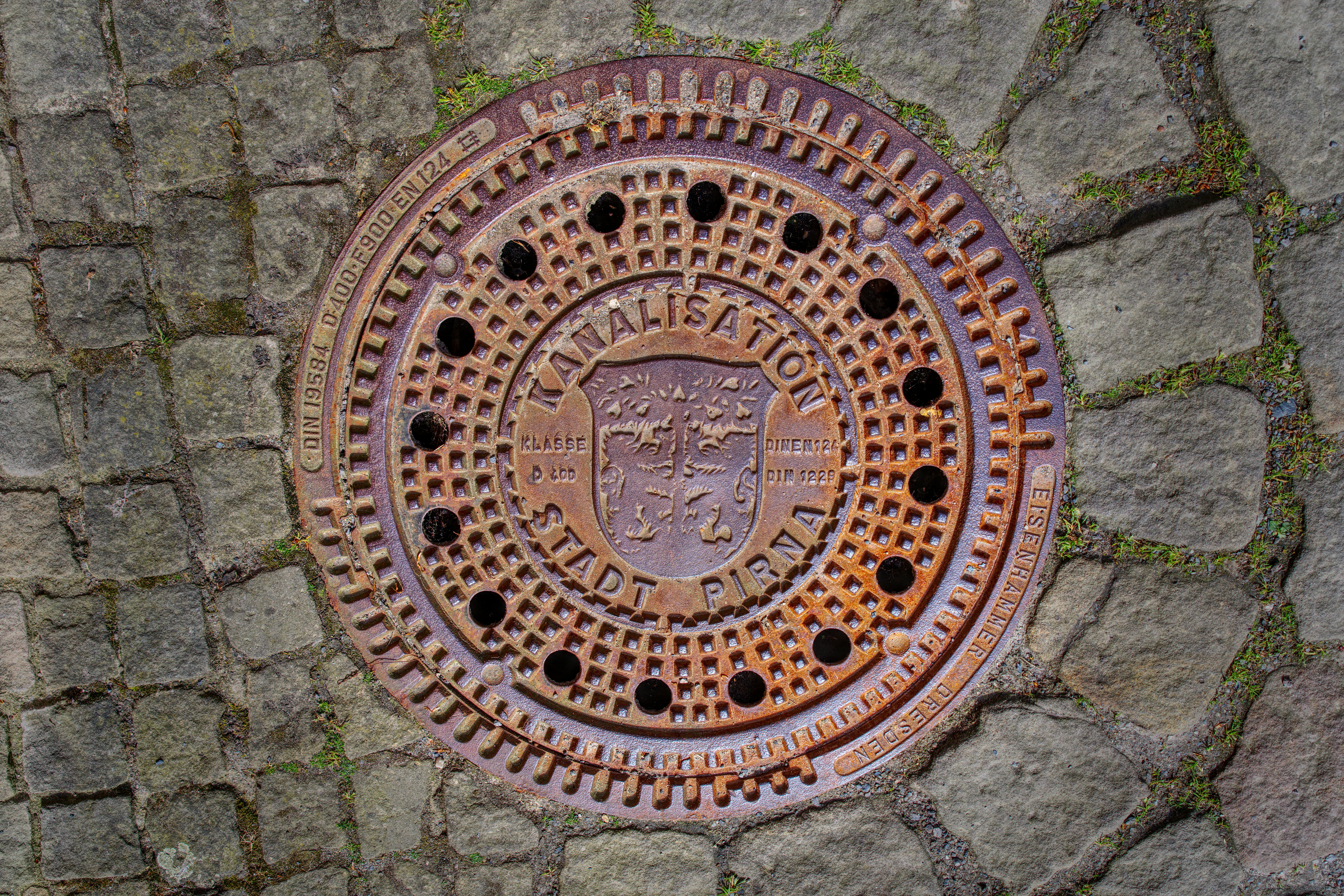 Intricately designed manhole cover featuring the inscription 'CANALISATION STADT PIENA,' set against a cobblestone background.