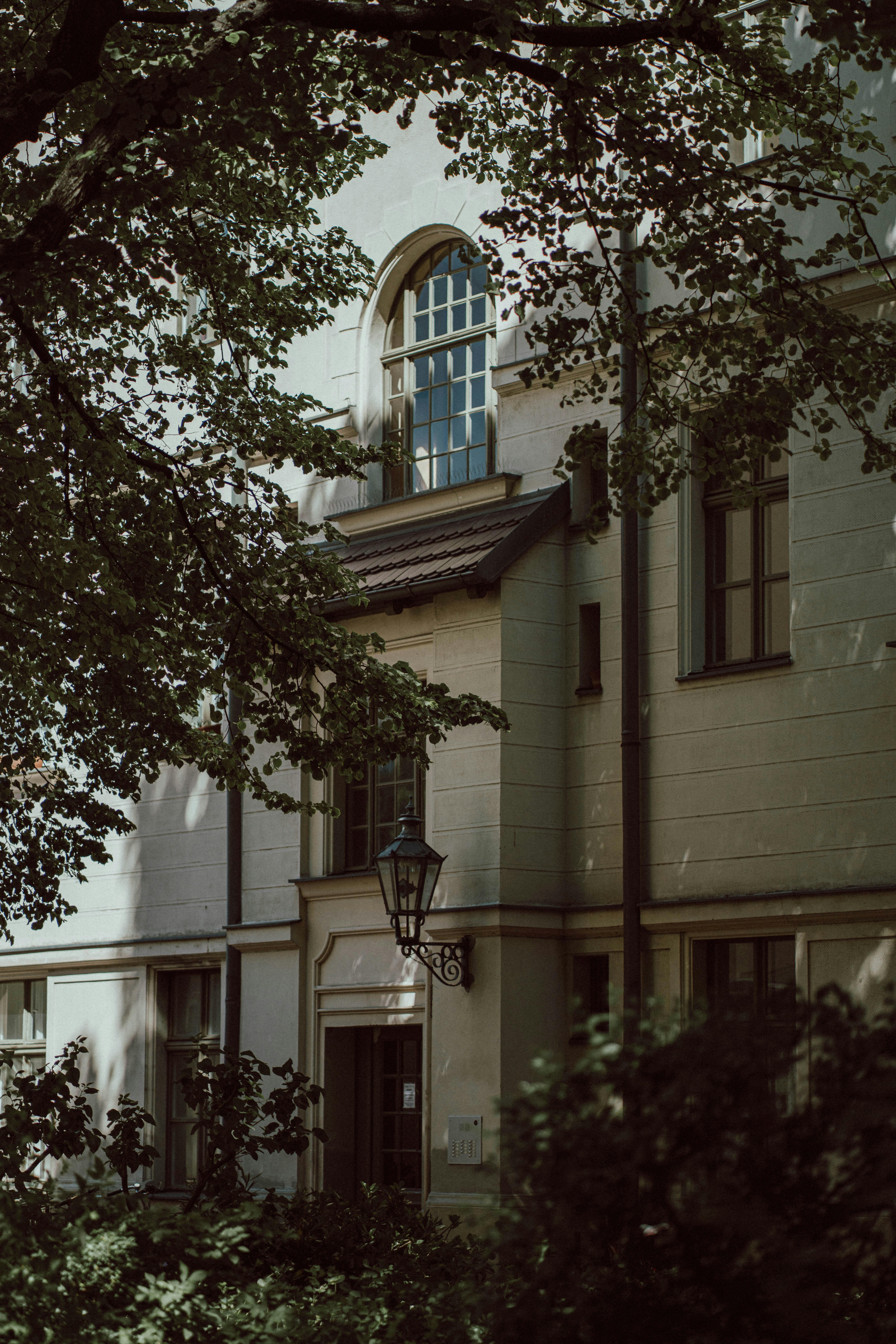Charming building facade framed by lush greenery and dappled sunlight, showcasing a vintage lantern and large windows.