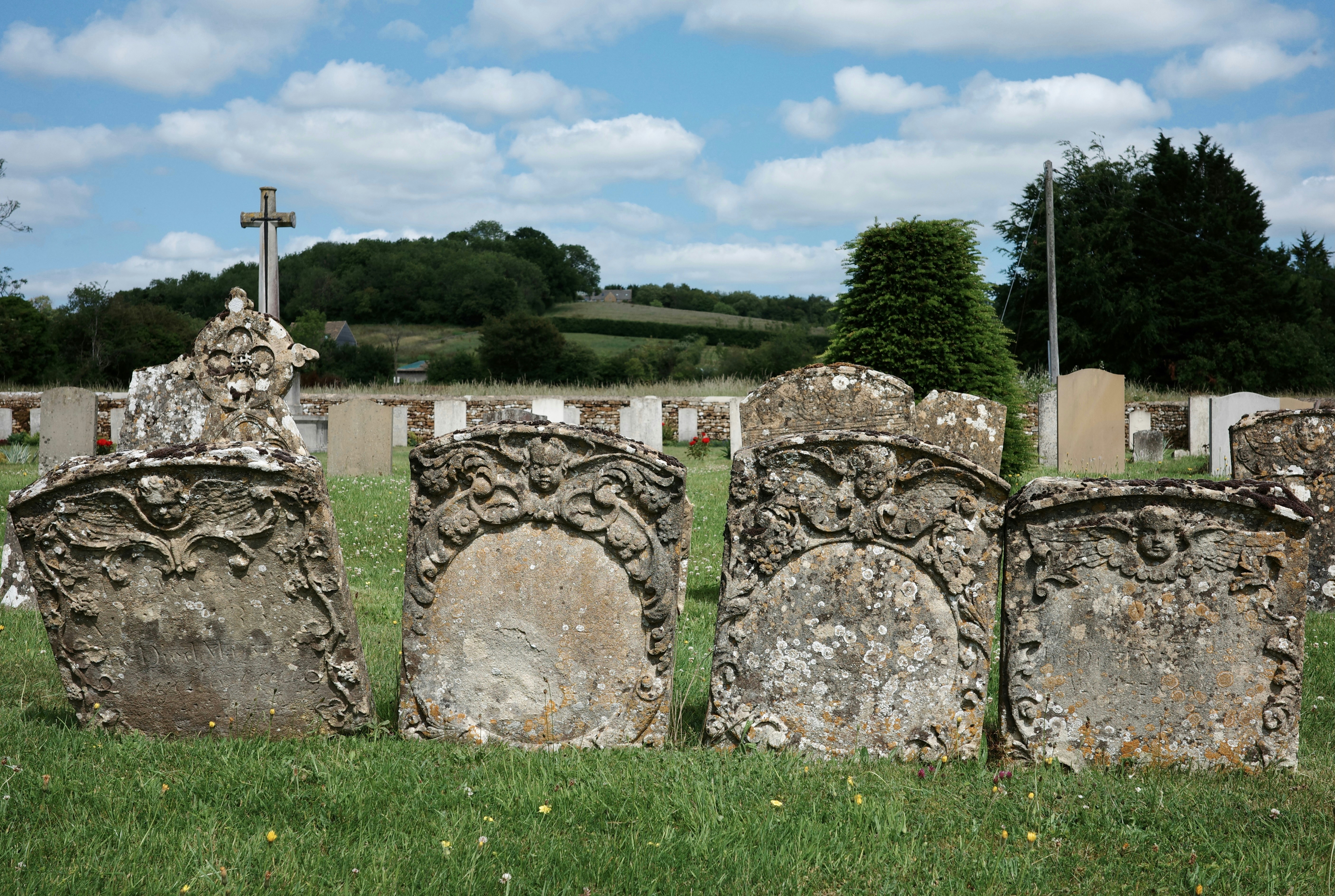 A row of weathered tombstones adorned with intricate carvings, set against a lush green cemetery backdrop under a bright blue sky.
