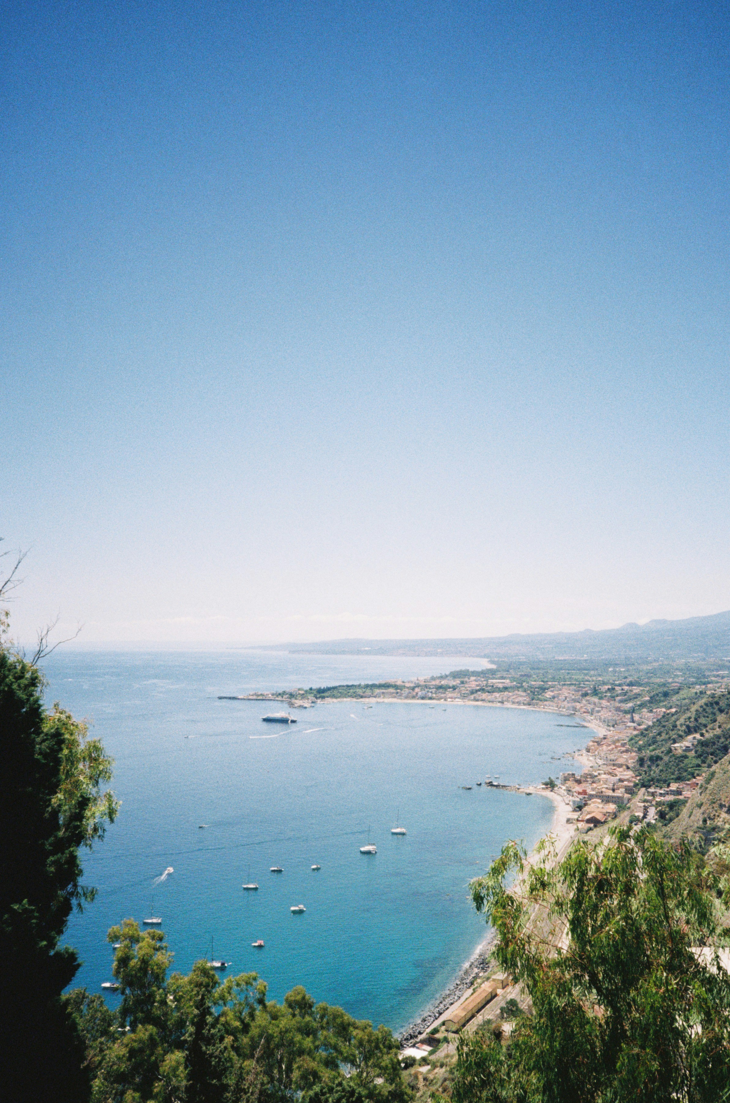 A panoramic view of a tranquil coastline with boats dotting the azure waters, framed by lush greenery and distant hills.
