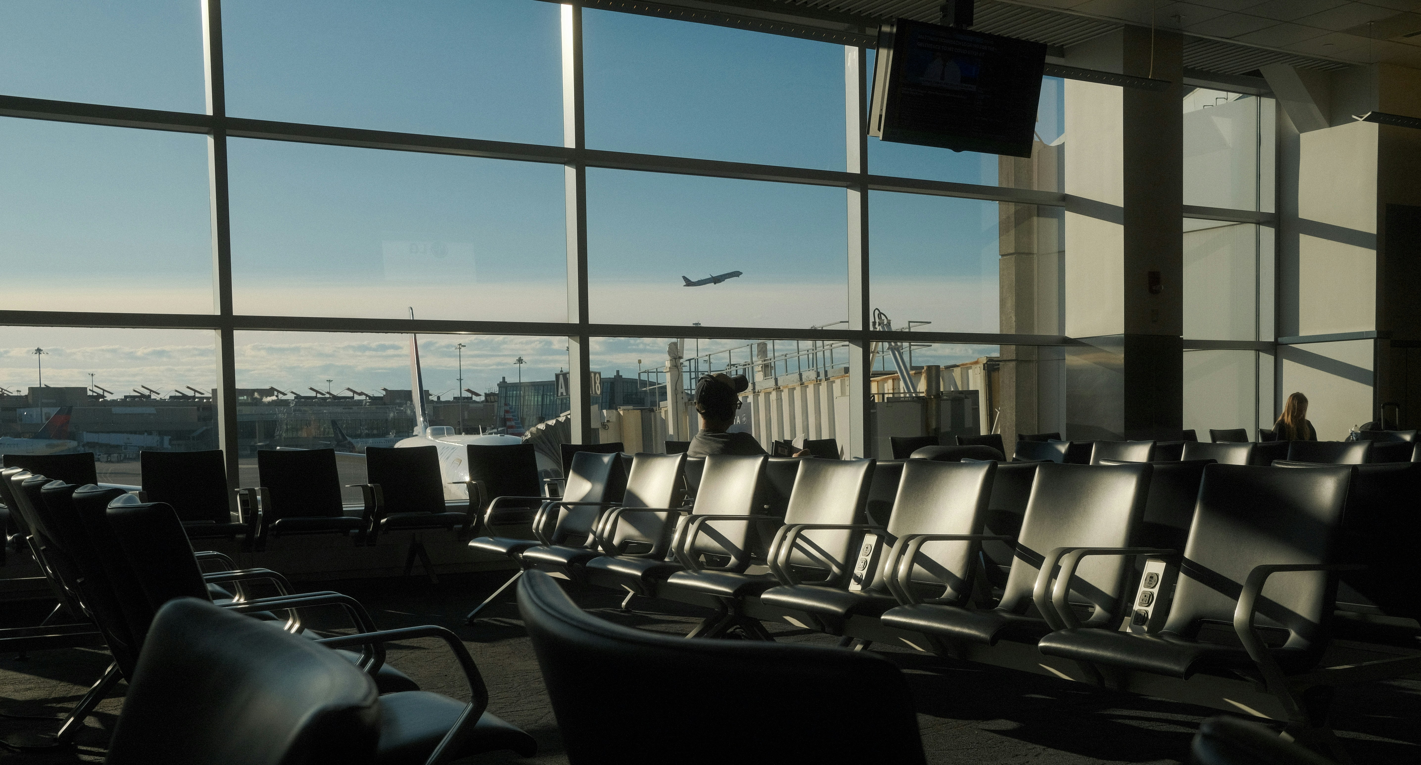 Empty airport terminal seats silhouetted against a bright sky, with a plane taking off in the distance.