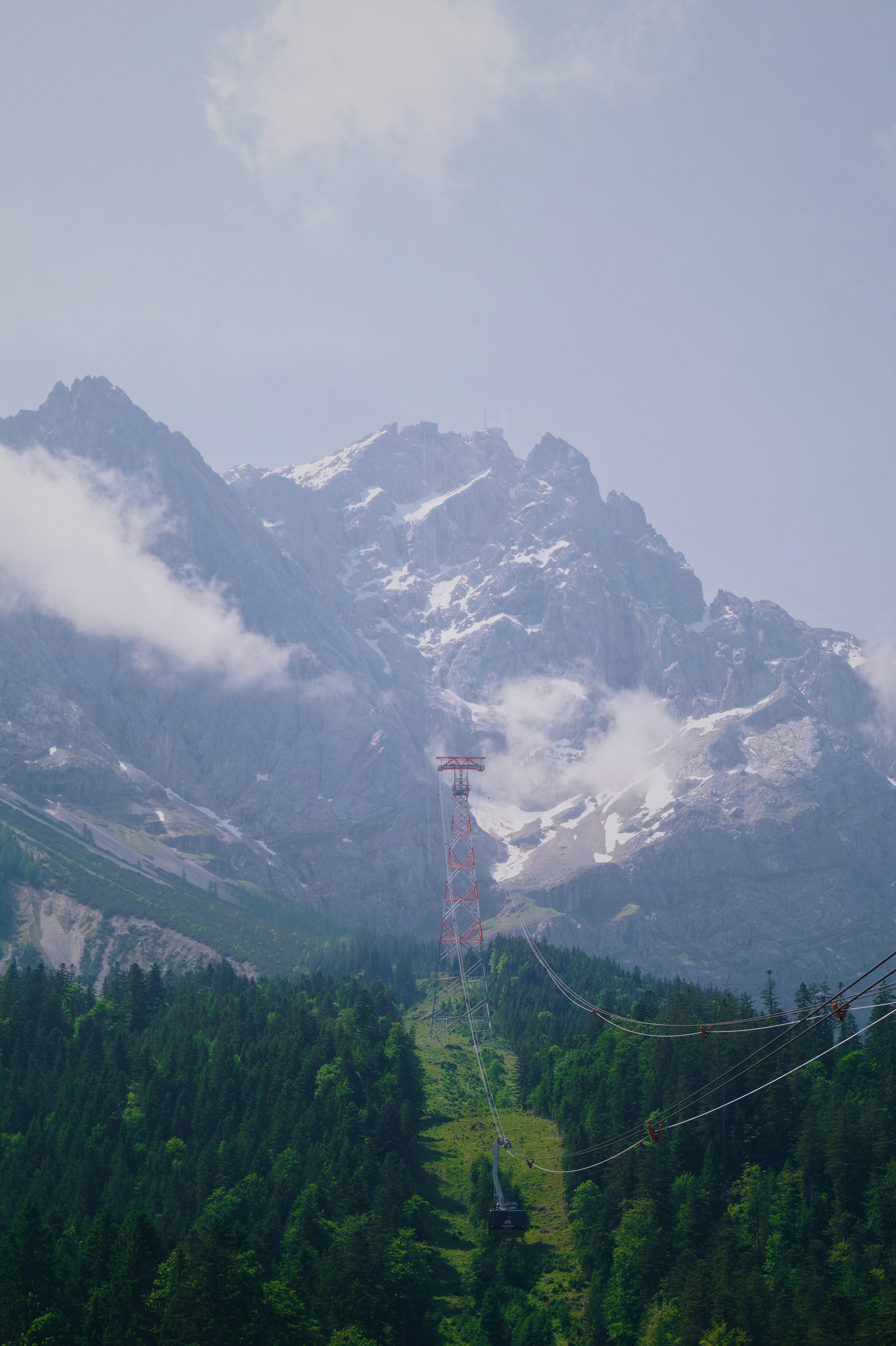 A cable car glides through lush green valleys towards majestic snow-capped peaks, framed by soft clouds.