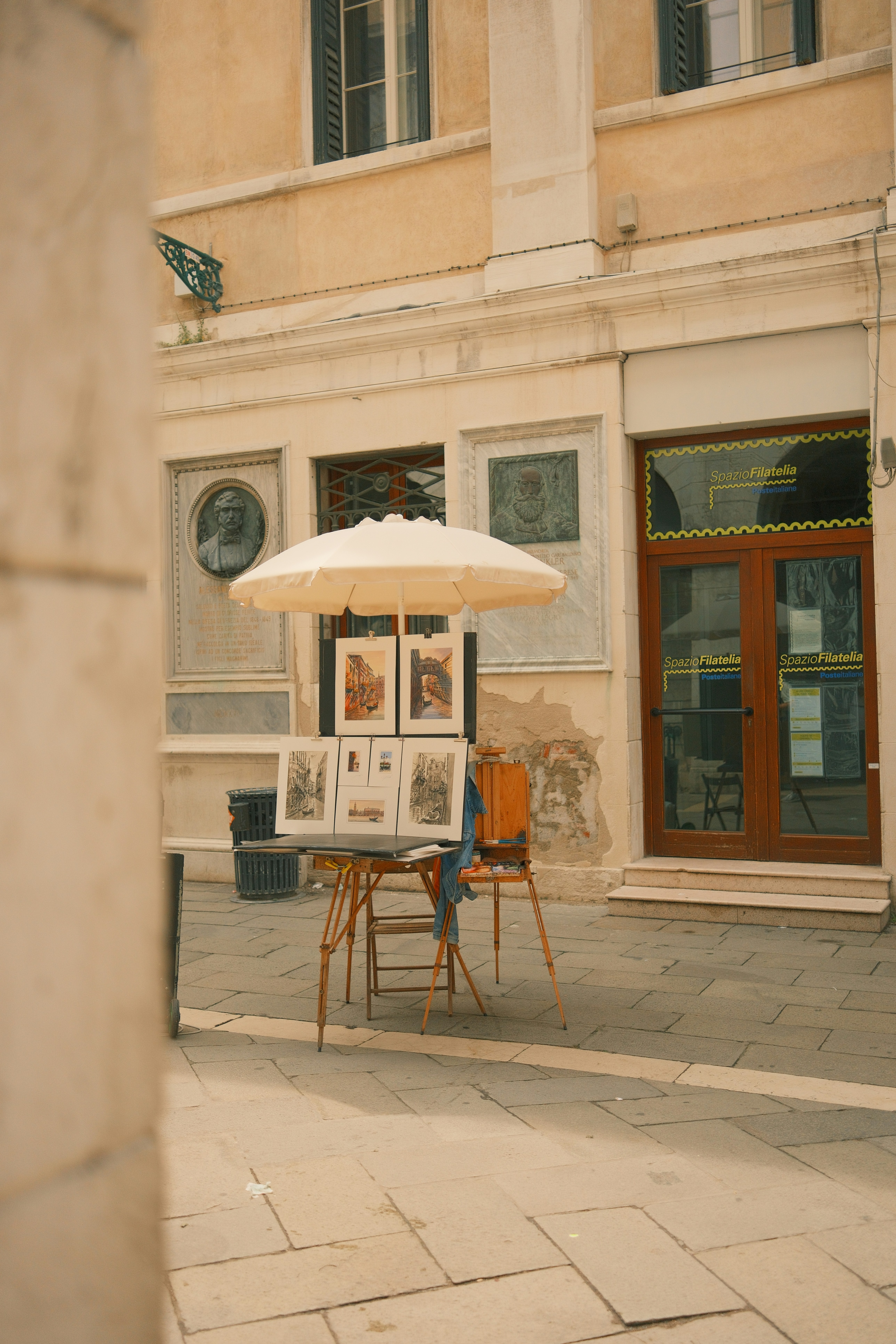 An outdoor art display featuring various framed artworks under a white umbrella, set against a charming historical backdrop.