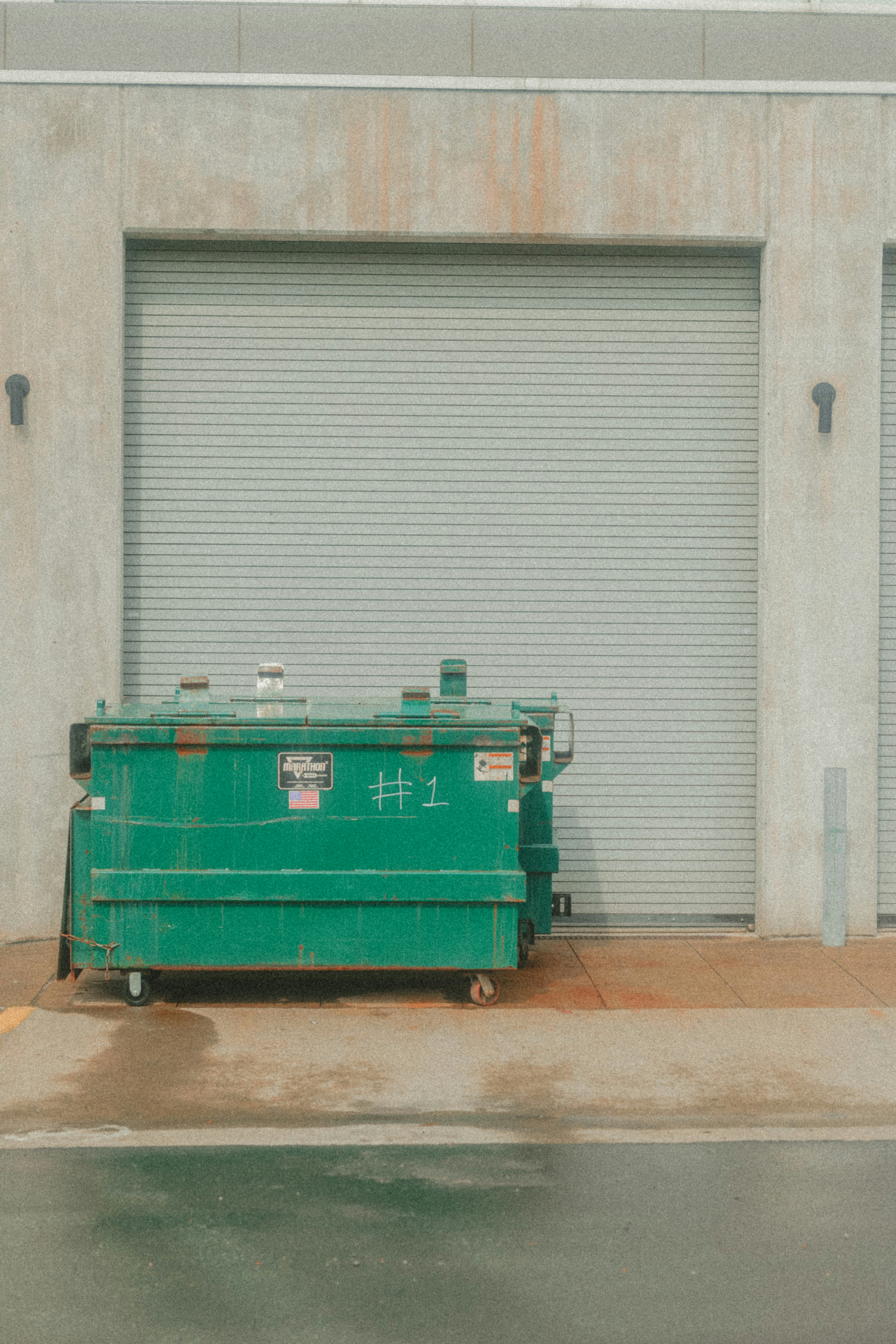Green dumpster positioned against a textured concrete wall with closed metal shutters, showcasing urban life’s overlooked details.