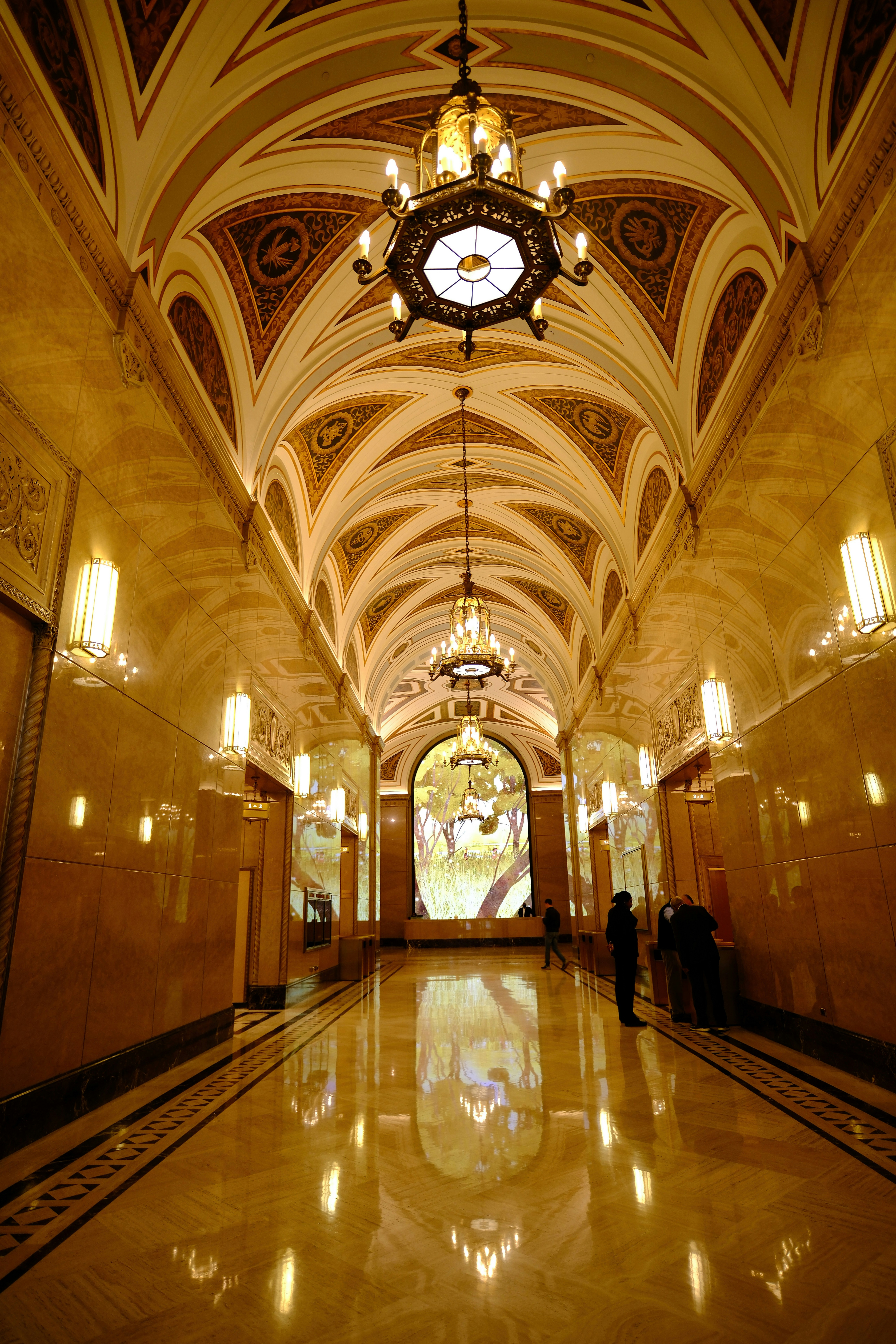 Luxurious hallway adorned with ornate chandeliers and intricate ceiling designs, reflecting a warm glow on polished marble floors.