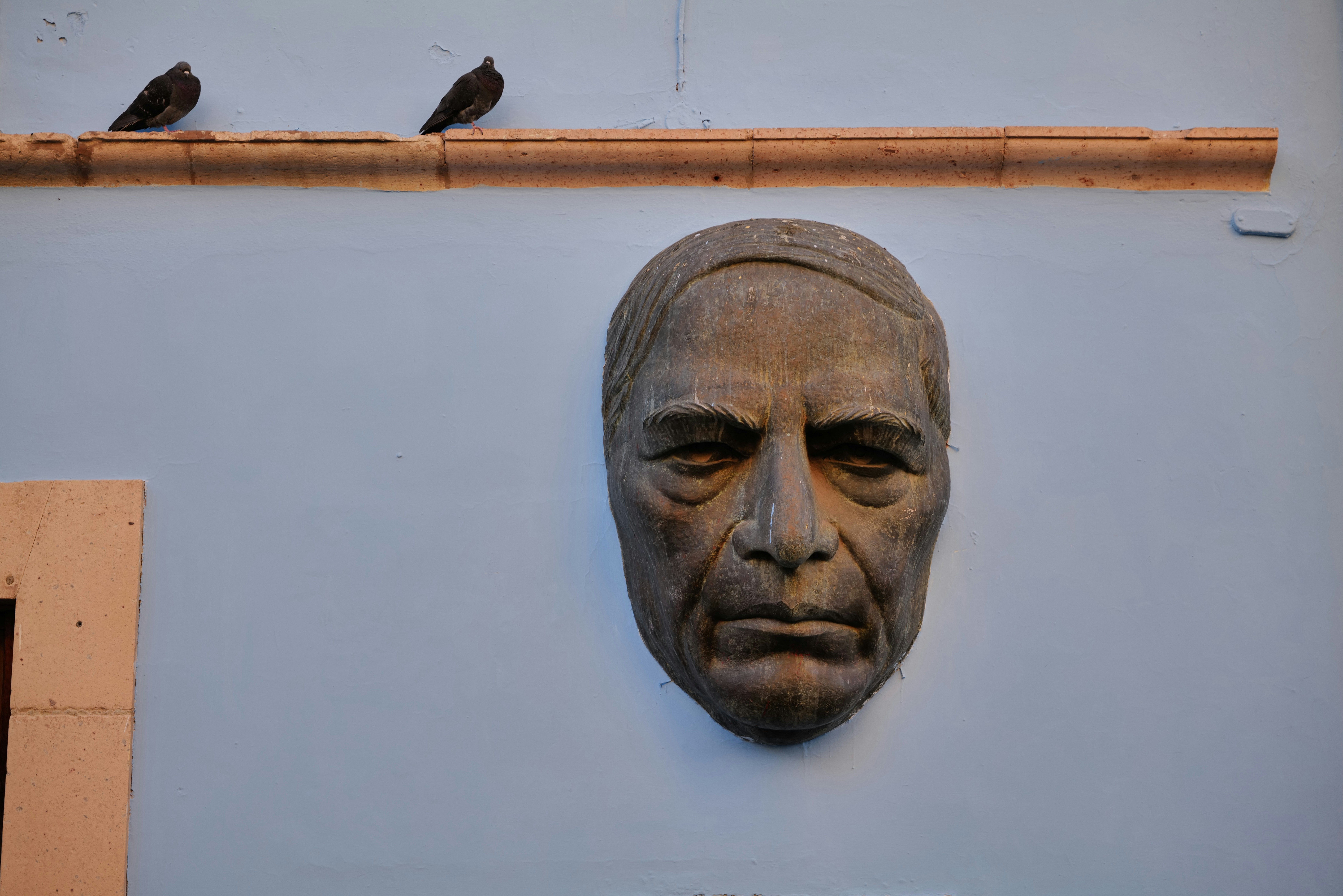 Cerulean Wall Portrait: A Weathered Mask Beneath a Perch of Birds