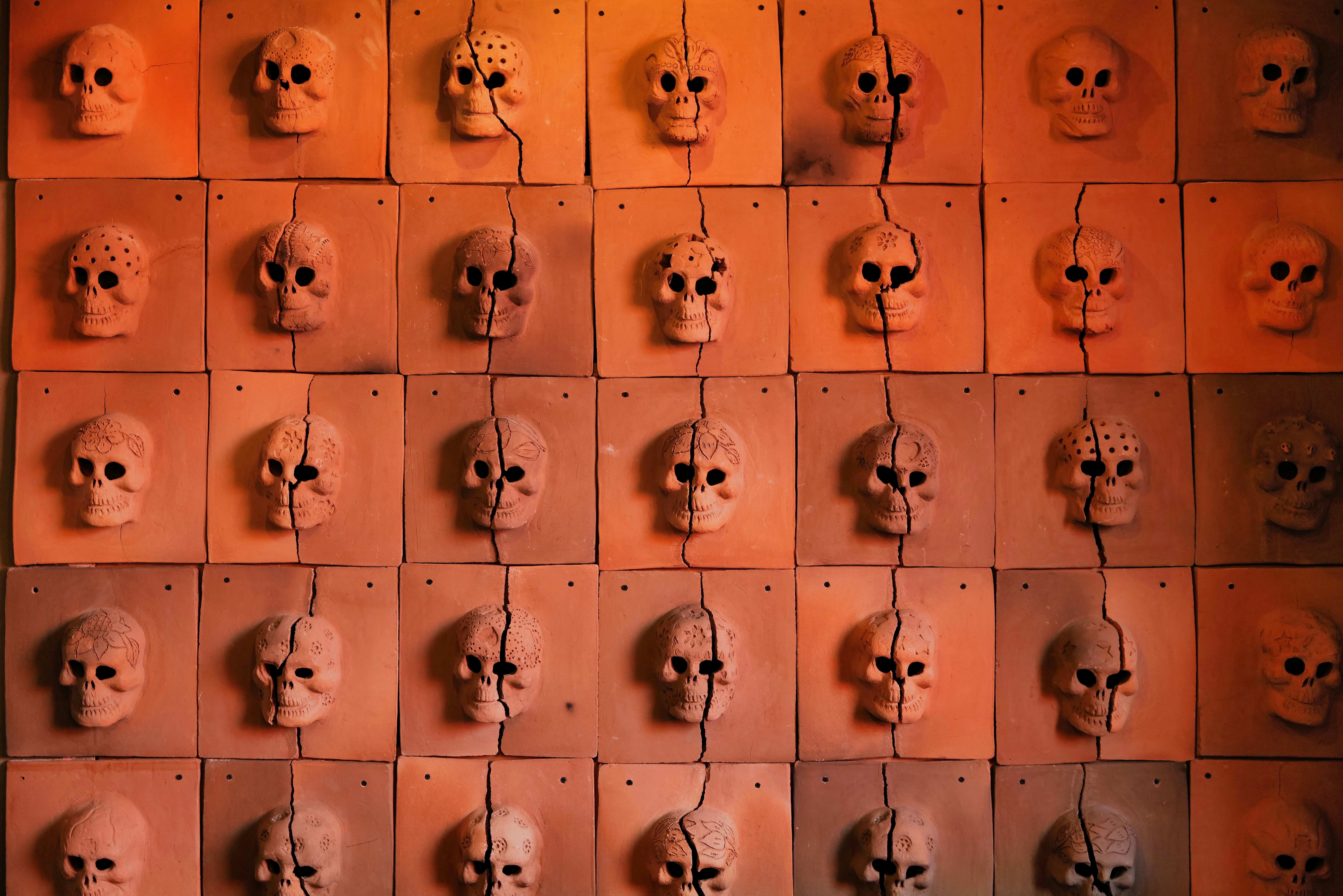 A grid of ceramic skulls, each uniquely marked and cracked, displayed against a warm, textured background. The arrangement invites contemplation on life and death.