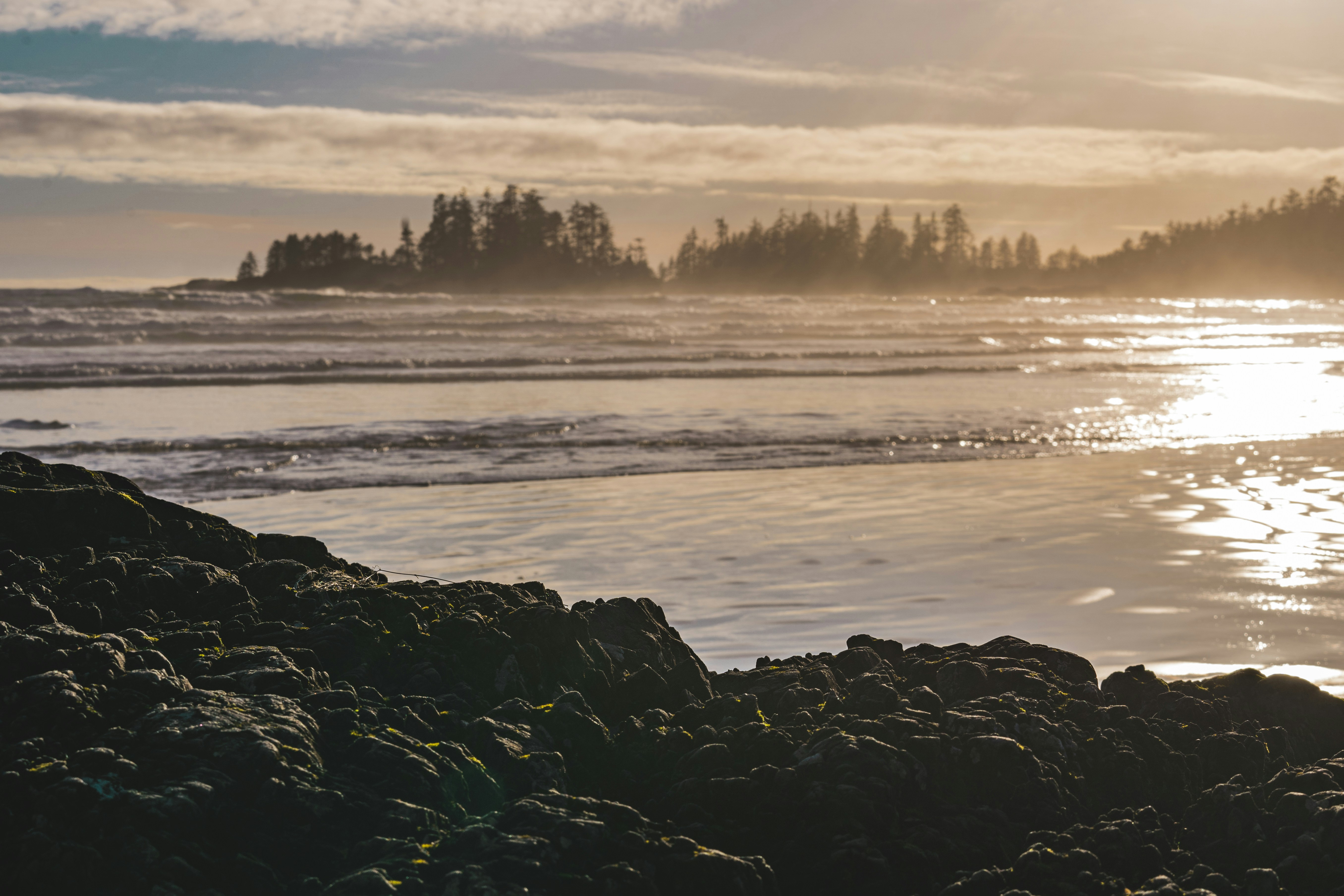 Gentle waves lap against rocky outcrops as mist envelops a distant island under a soft, golden light.