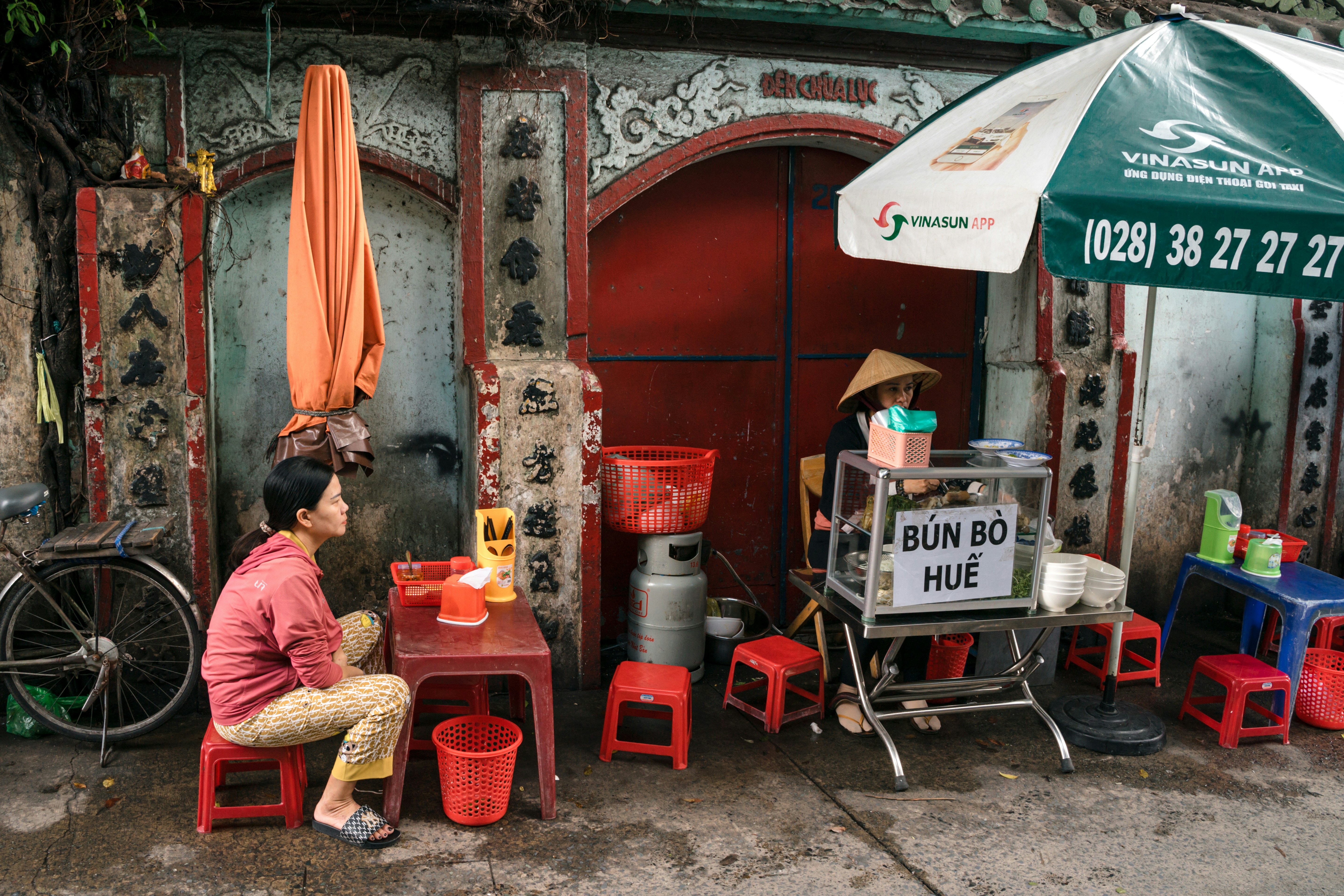 A candid street food scene in District 4, Saigon, Vietnam. A woman waits for her bowl of bún bò Huế at a small roadside stall set against the weathered facade of an old temple. The red stools, umbrellas, and layered textures reflect the character of daily life in Vietnam.
