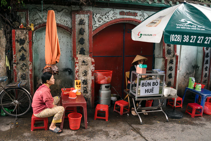 A woman sits on a red plastic stool beside a small street food cart labeled "Bún Bò Huế," under a green-and-white umbrella. Old painted walls, stacked stools, and a parked bicycle frame the narrow roadside scene.