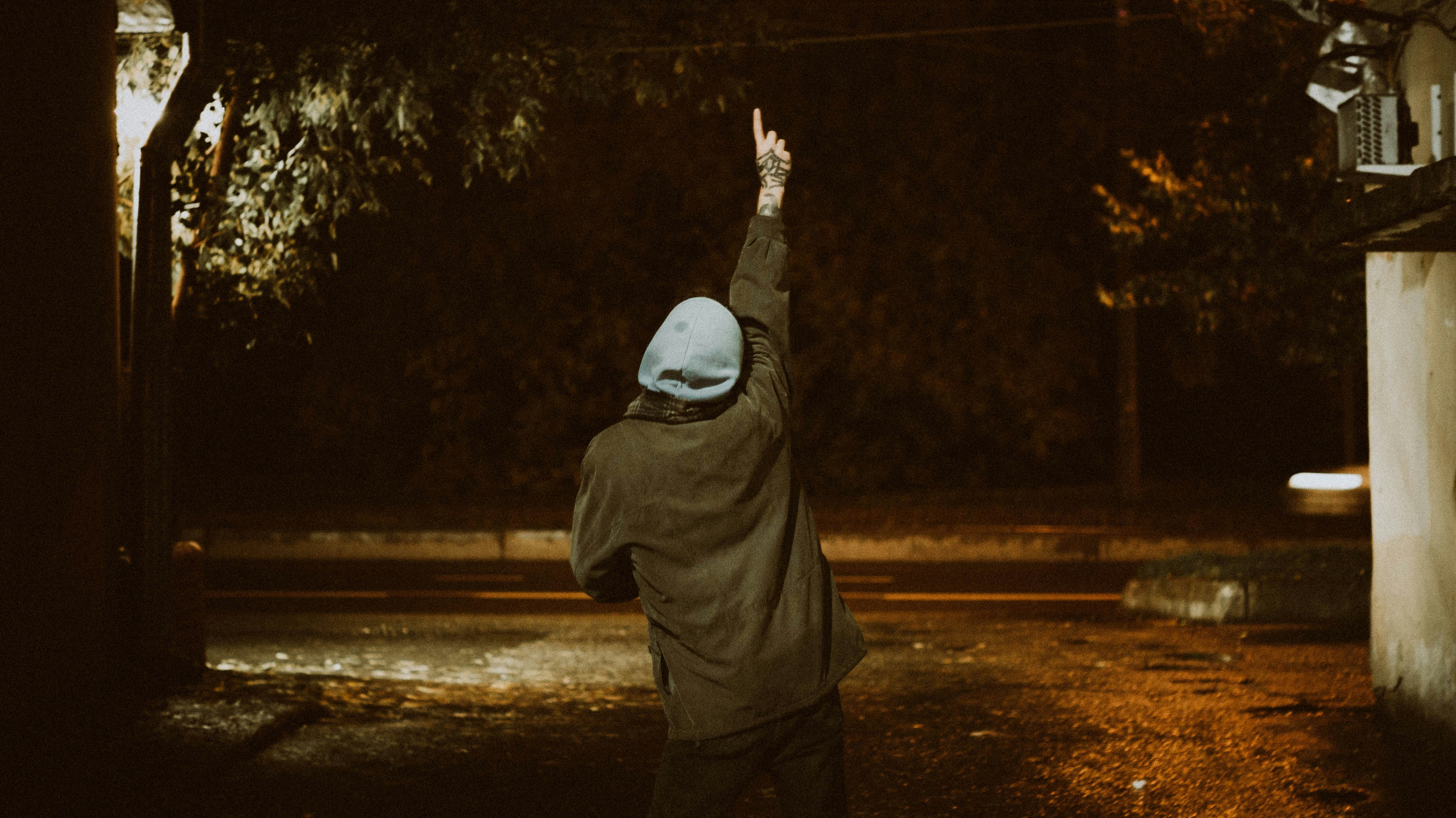 A figure in a hoodie gestures towards the night sky, illuminated by ambient street lighting. The scene captures a moment of reflection and solitude.