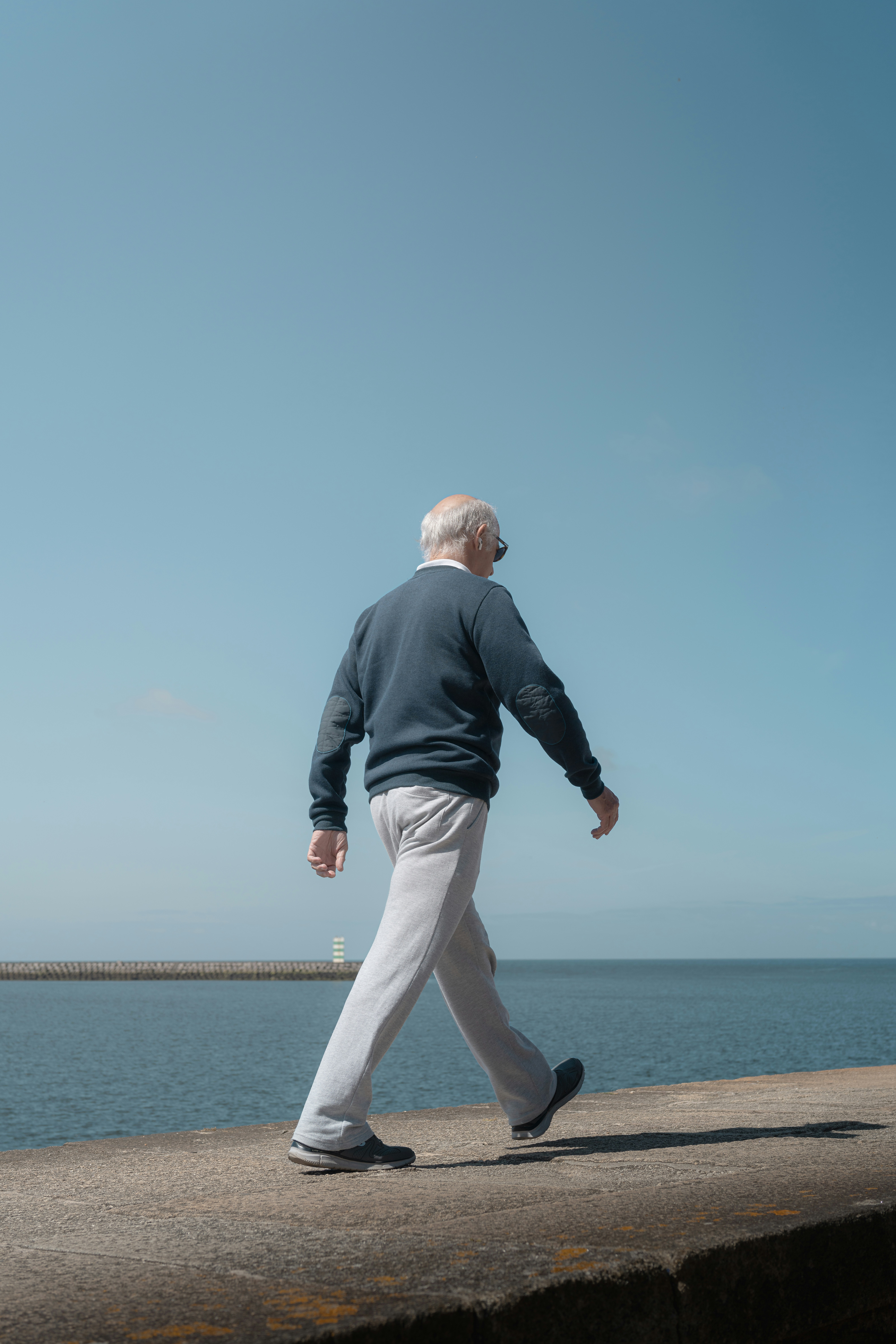 Elderly man strolling along a coastal path, with a clear blue sky and calm sea in the background.