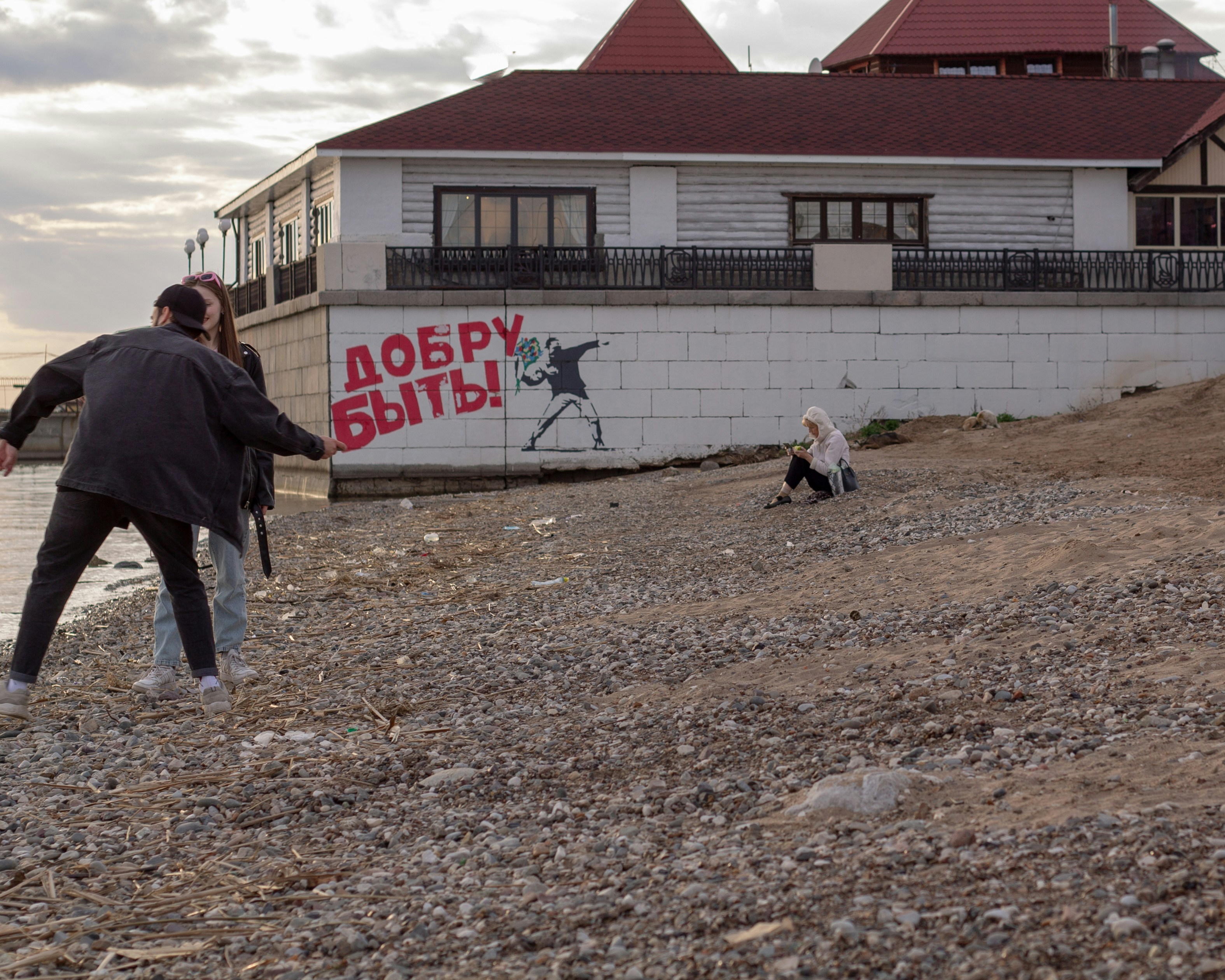 Two figures interact playfully on a pebbled beach near a building adorned with a striking mural. A lone figure sits nearby, immersed in thought.