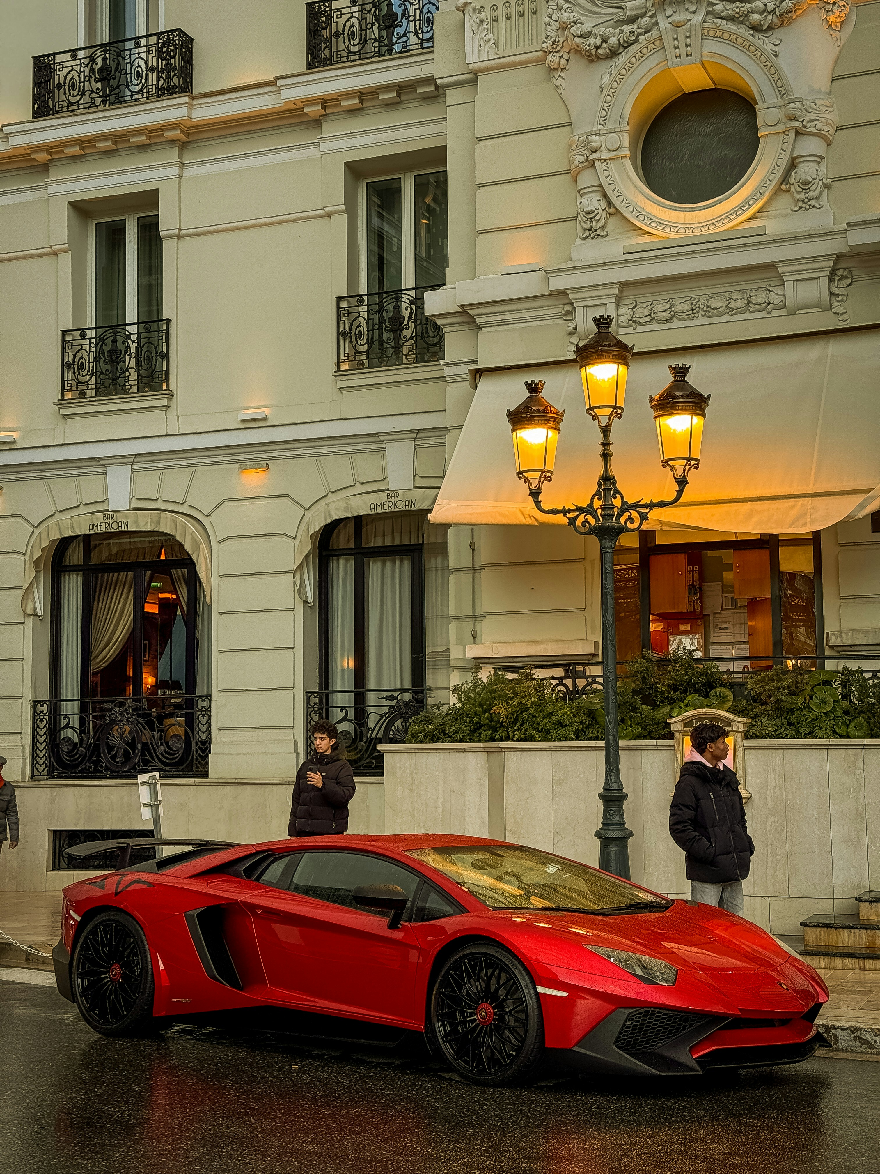 A striking red sports car parked under ornate street lamps outside an elegant building, showcasing a blend of luxury and urban life.