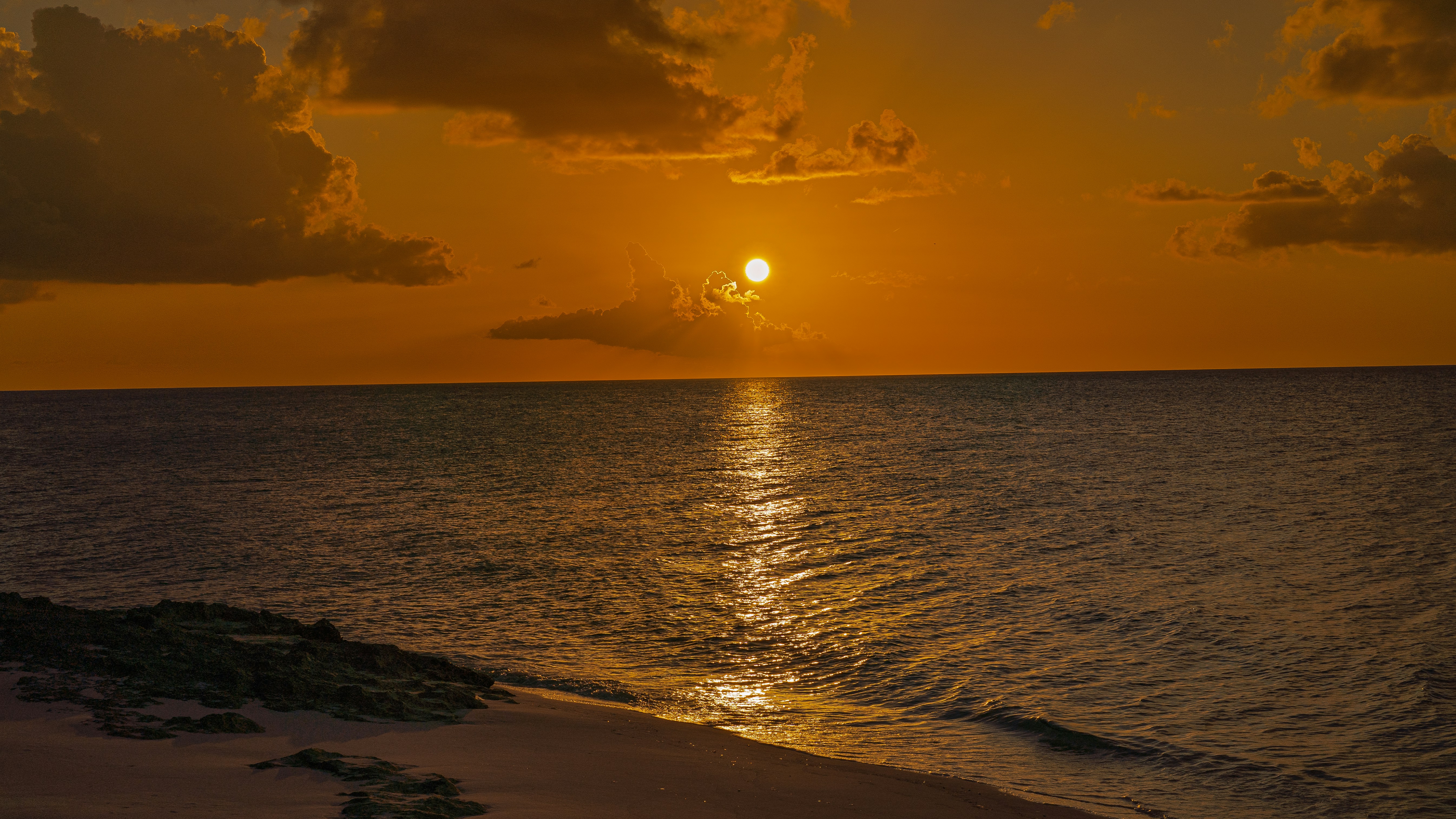 Golden sunset casting warm hues over calm ocean waters, with reflections shimmering on the surface. Clouds softly frame the sun as it descends.
