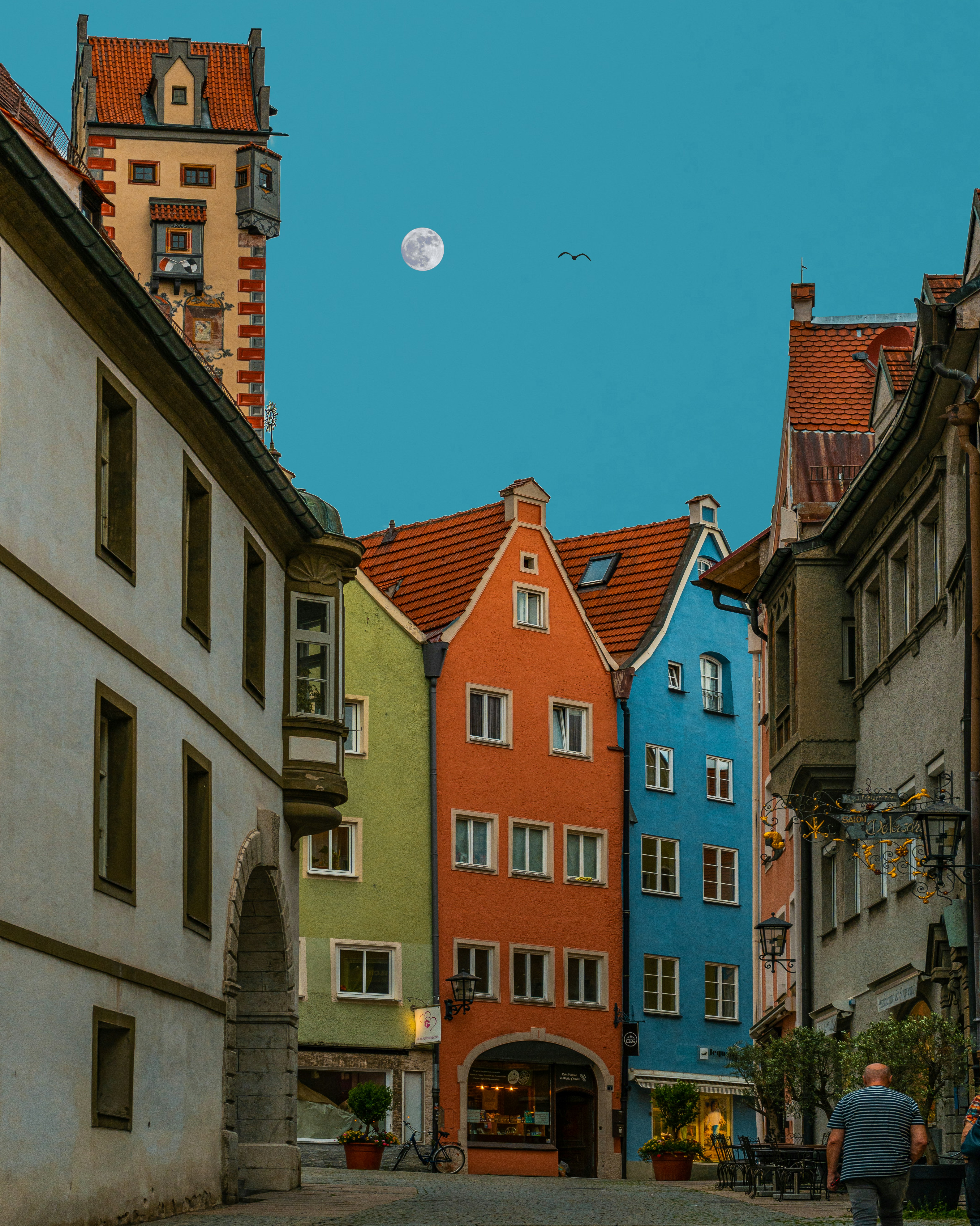 Charming alleyway adorned with colorful buildings under a clear blue sky, featuring a bright moon and a solitary figure walking. A tranquil scene capturing the essence of urban life.