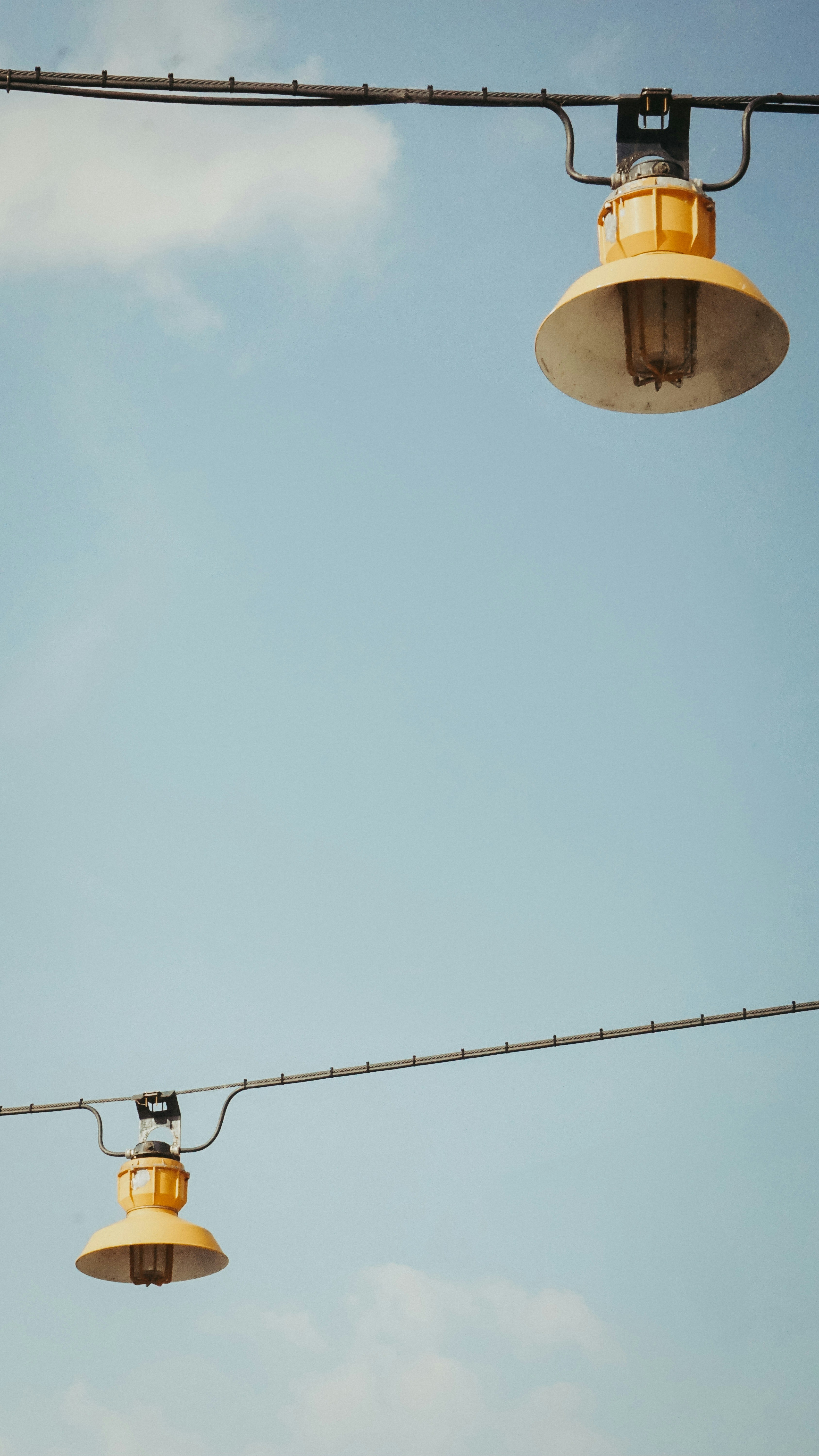 Two yellow hanging lamps suspended above a clear blue sky, casting a warm glow. The simplicity of the scene highlights the interplay between light and space.