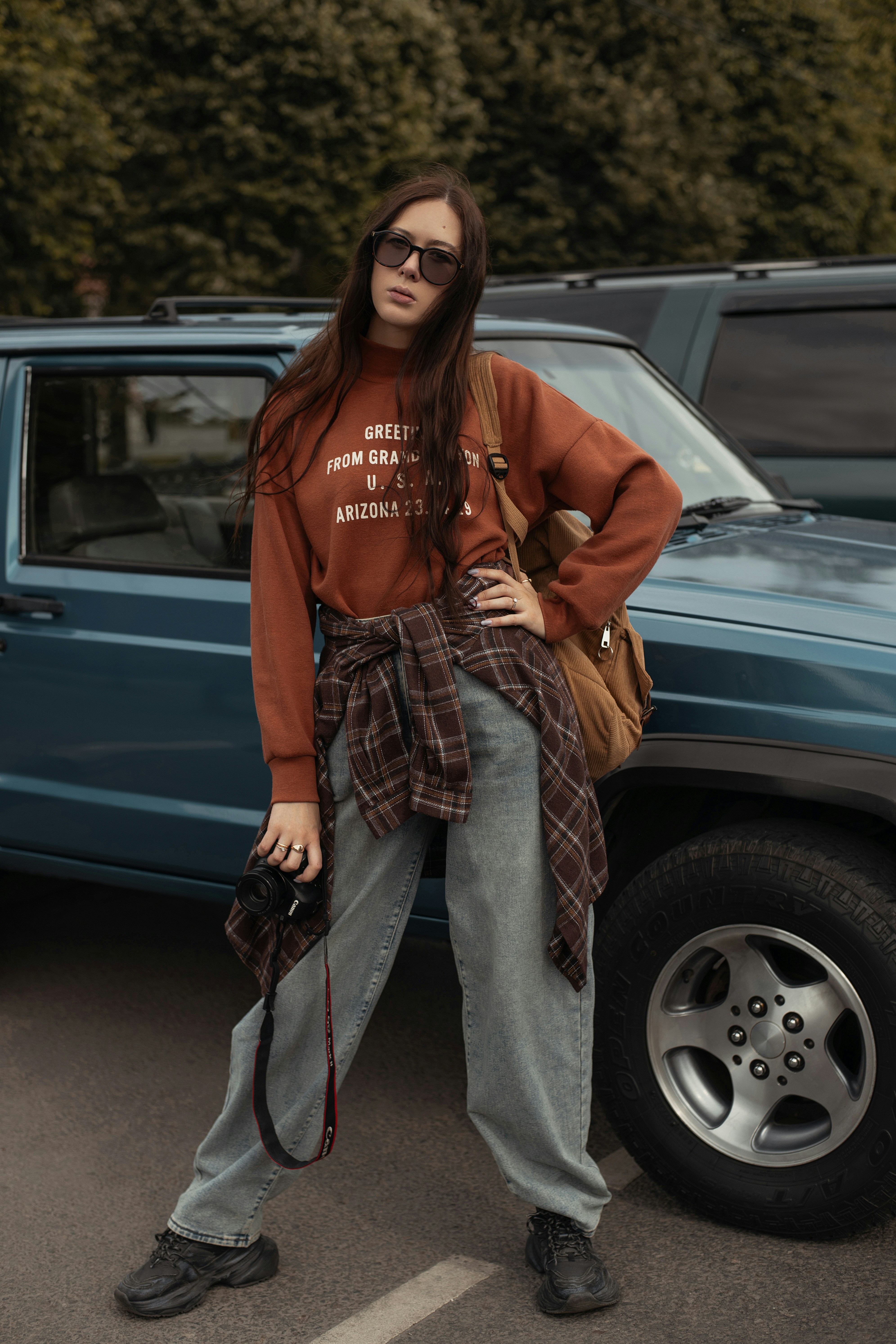Young woman in casual attire poses confidently beside a vintage blue SUV, showcasing a blend of retro and contemporary fashion. Her stylish accessories complement the urban setting.