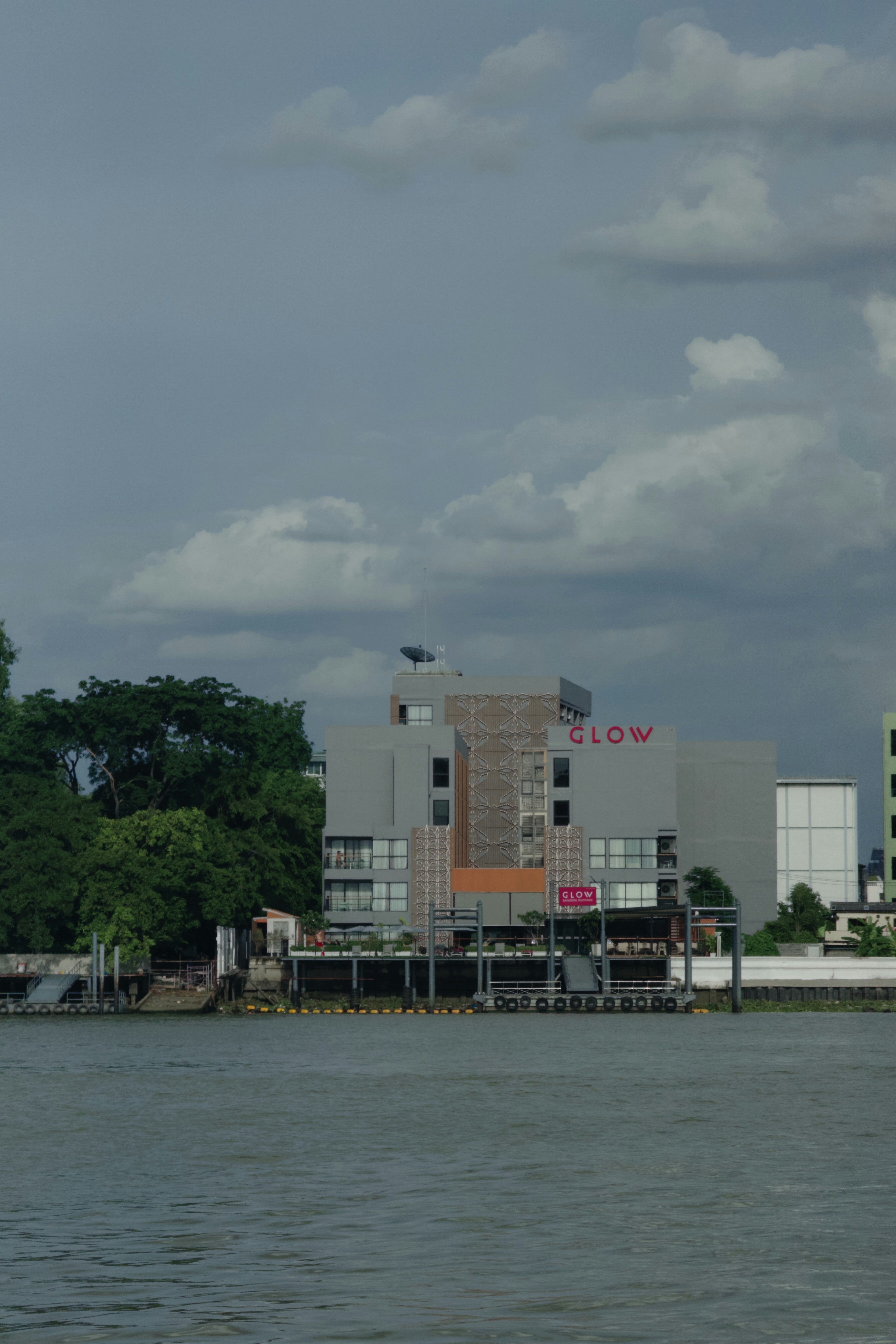 Modern building with 'GLOW' signage beside a river, framed by lush greenery and a dramatic sky.