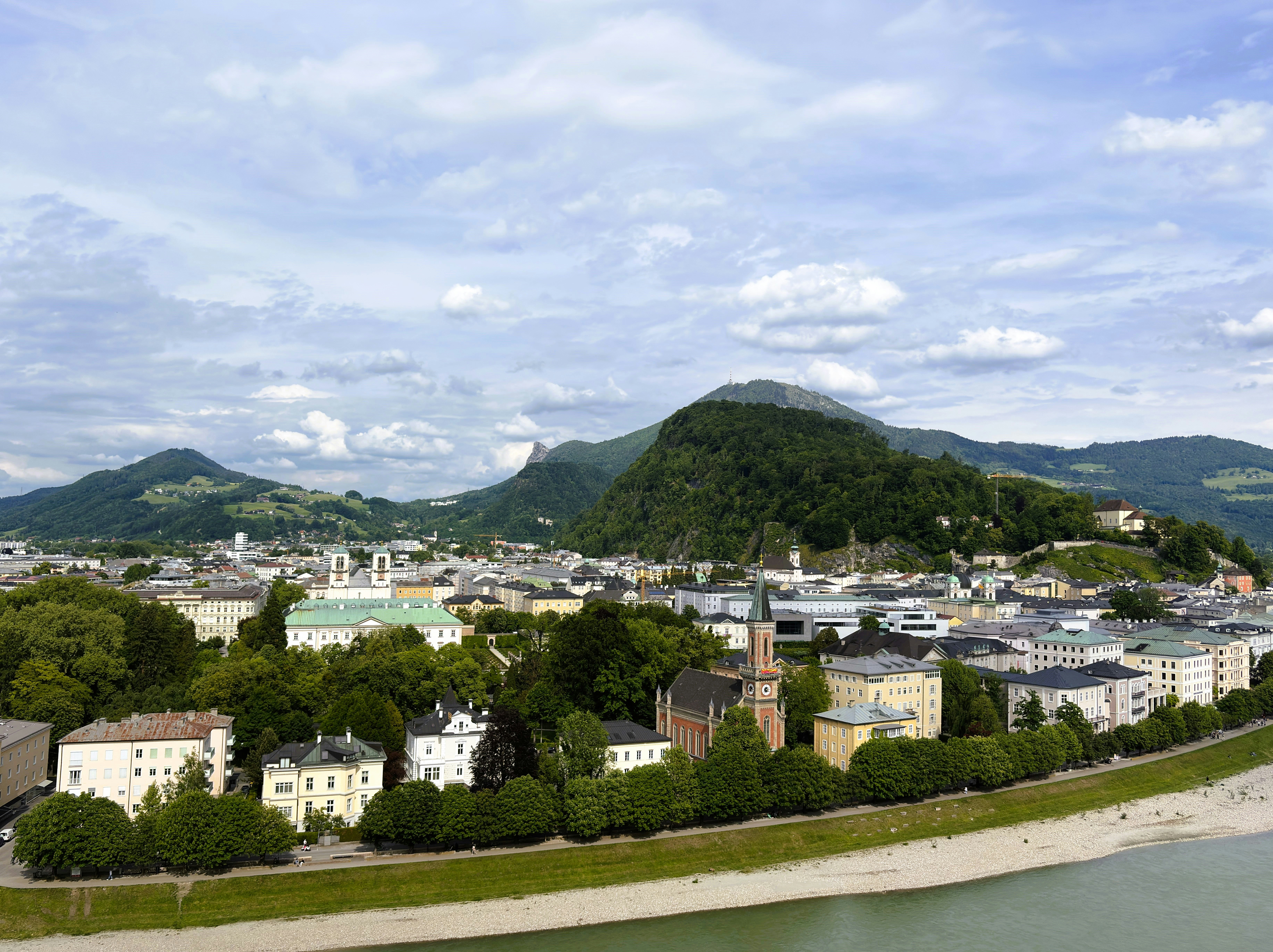 Panoramic view of Salzburg featuring lush greenery, historic architecture, and the distant mountains. The image showcases the city's blend of nature and urban life.