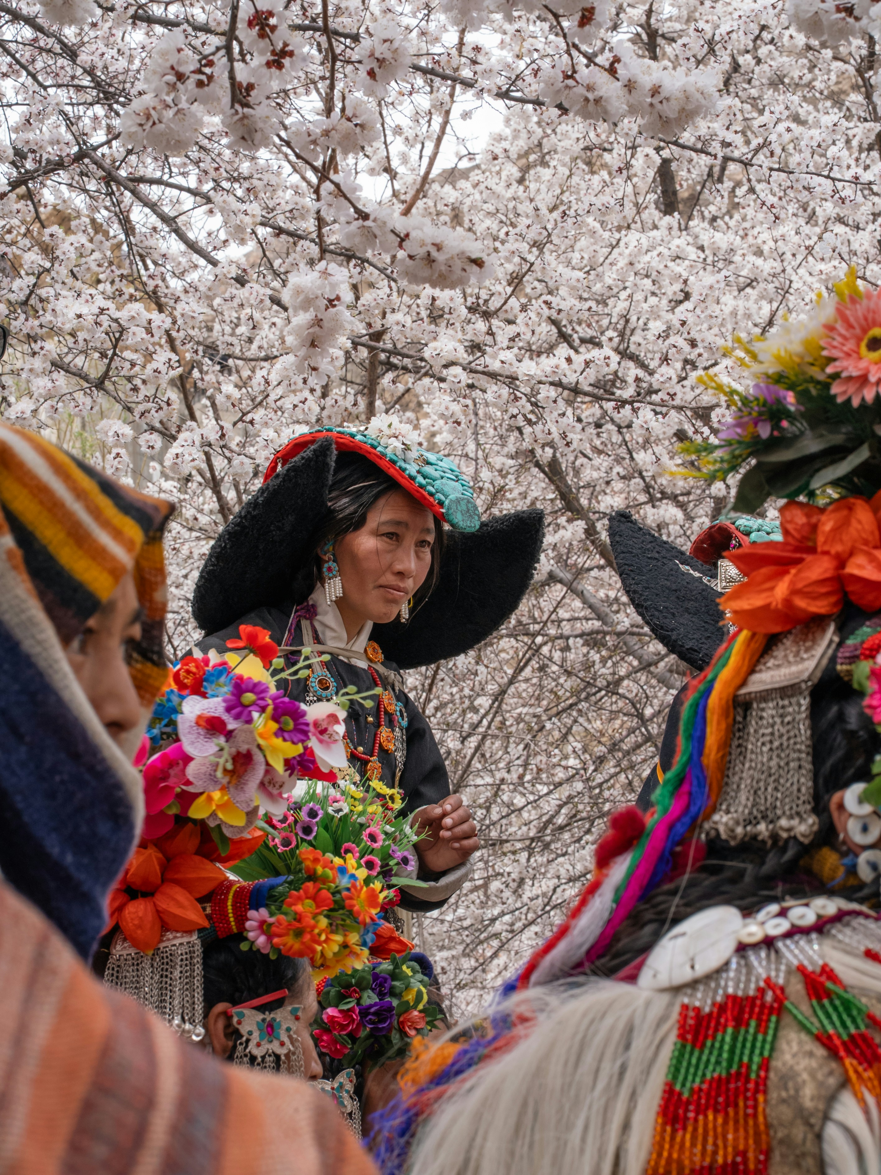 A woman adorned in vibrant traditional attire holds a bouquet of flowers, surrounded by cherry blossoms in full bloom.