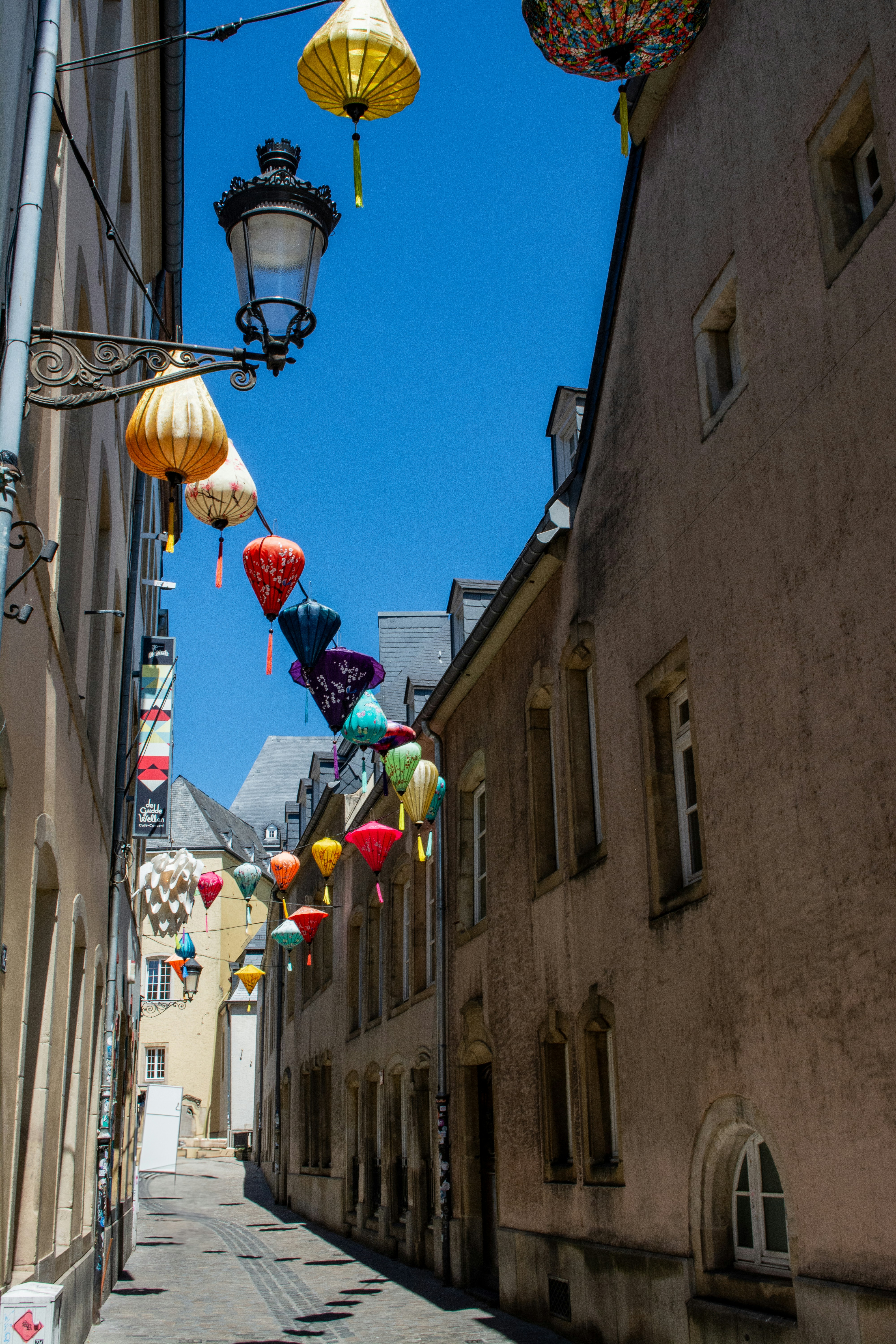 A narrow cobblestone street in Luxembourg comes alive with a canopy of colorful paper lanterns strung between historic buildings, casting playful shadows under a radiant blue sky. The vibrant display adds a festive charm to the quaint alleyway, merging tradition with artistic flair.