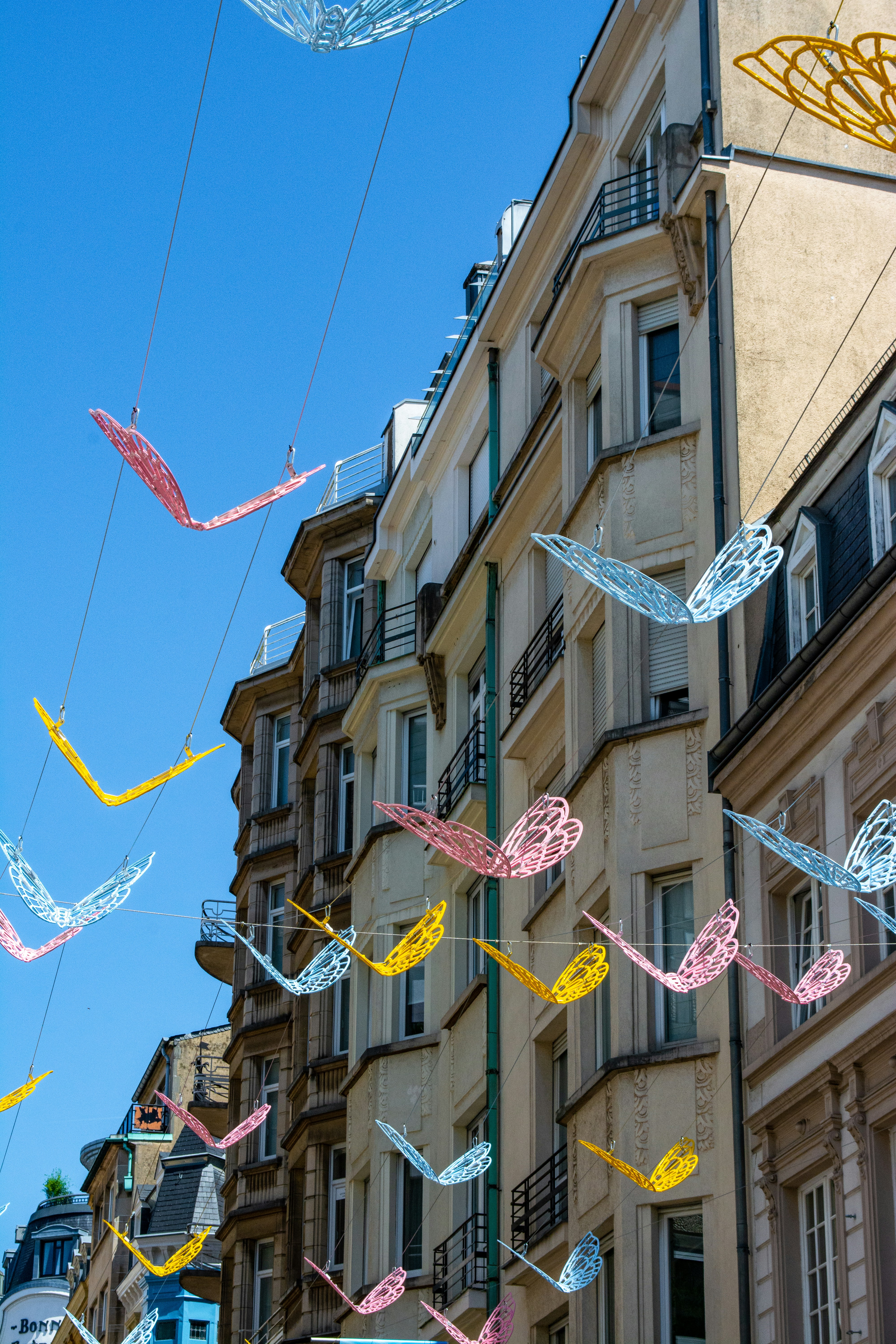 Colorful paper birds suspended between buildings against a clear blue sky, creating a playful urban installation.