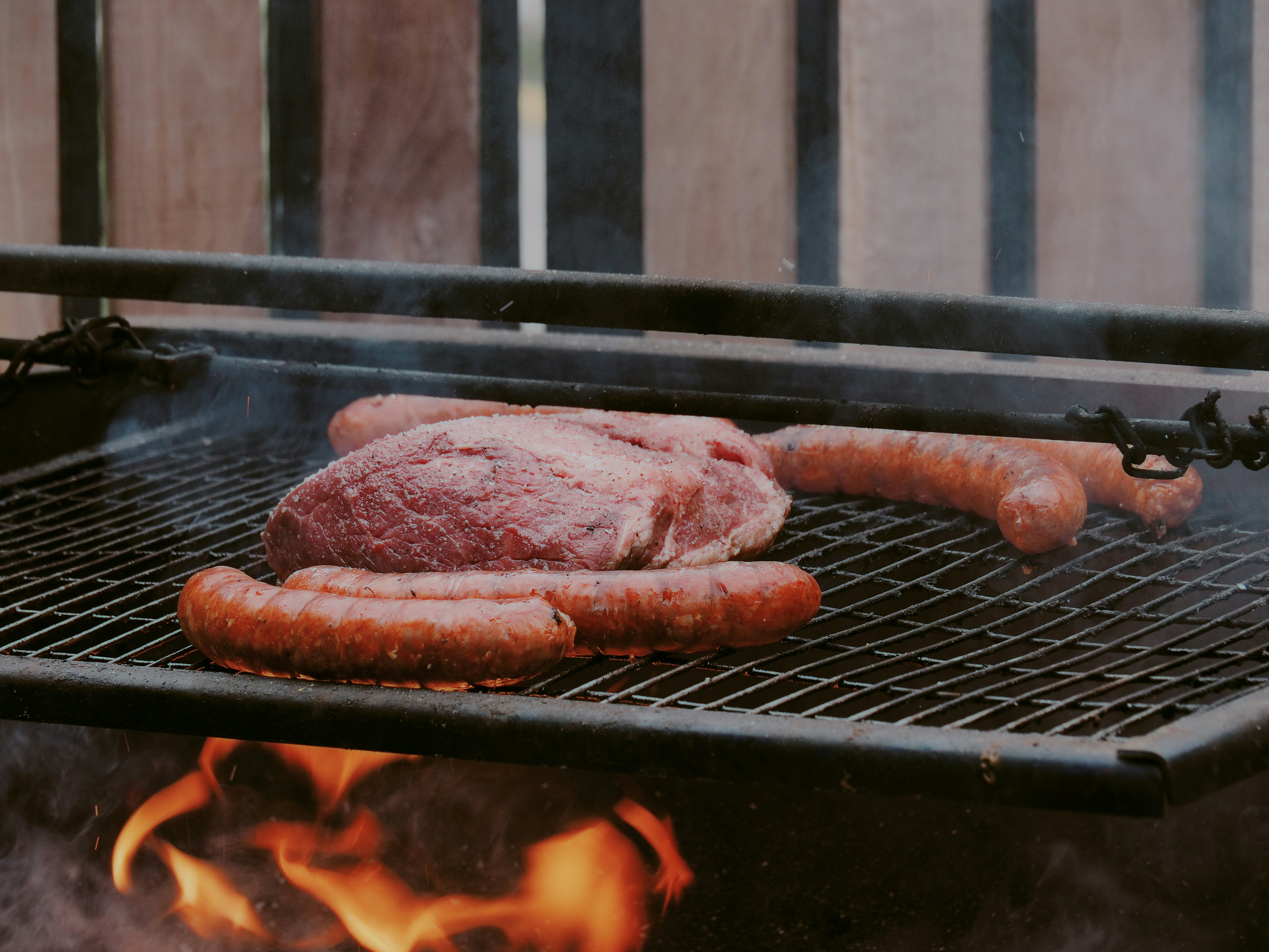 Raw meats including sausages and a large cut of beef on a grill, with flames and smoke enhancing the cooking atmosphere.