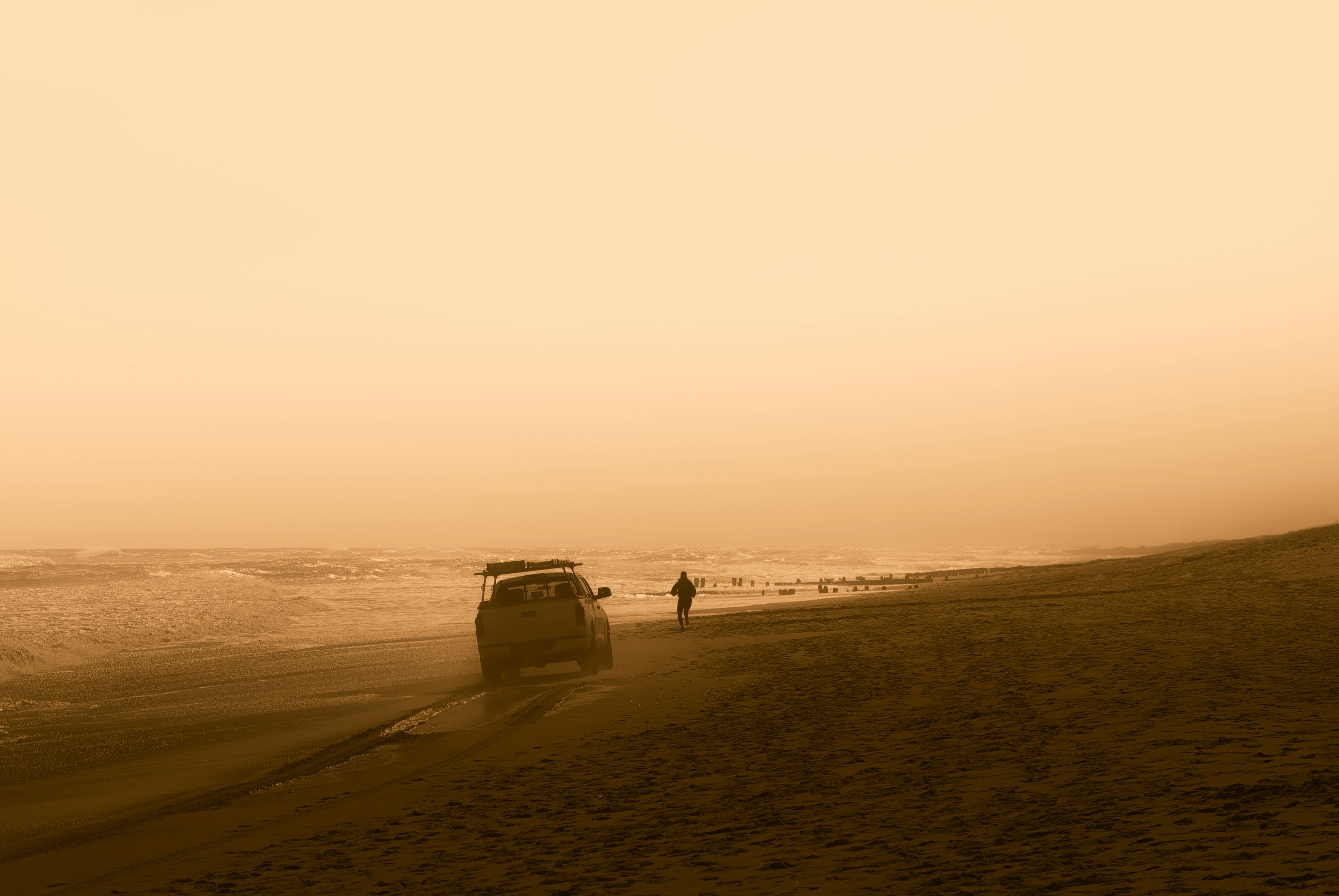 A lone figure walks along a misty beach, with a vehicle parked nearby, embodying a sense of quiet reflection. The sepia tones enhance the tranquil atmosphere.