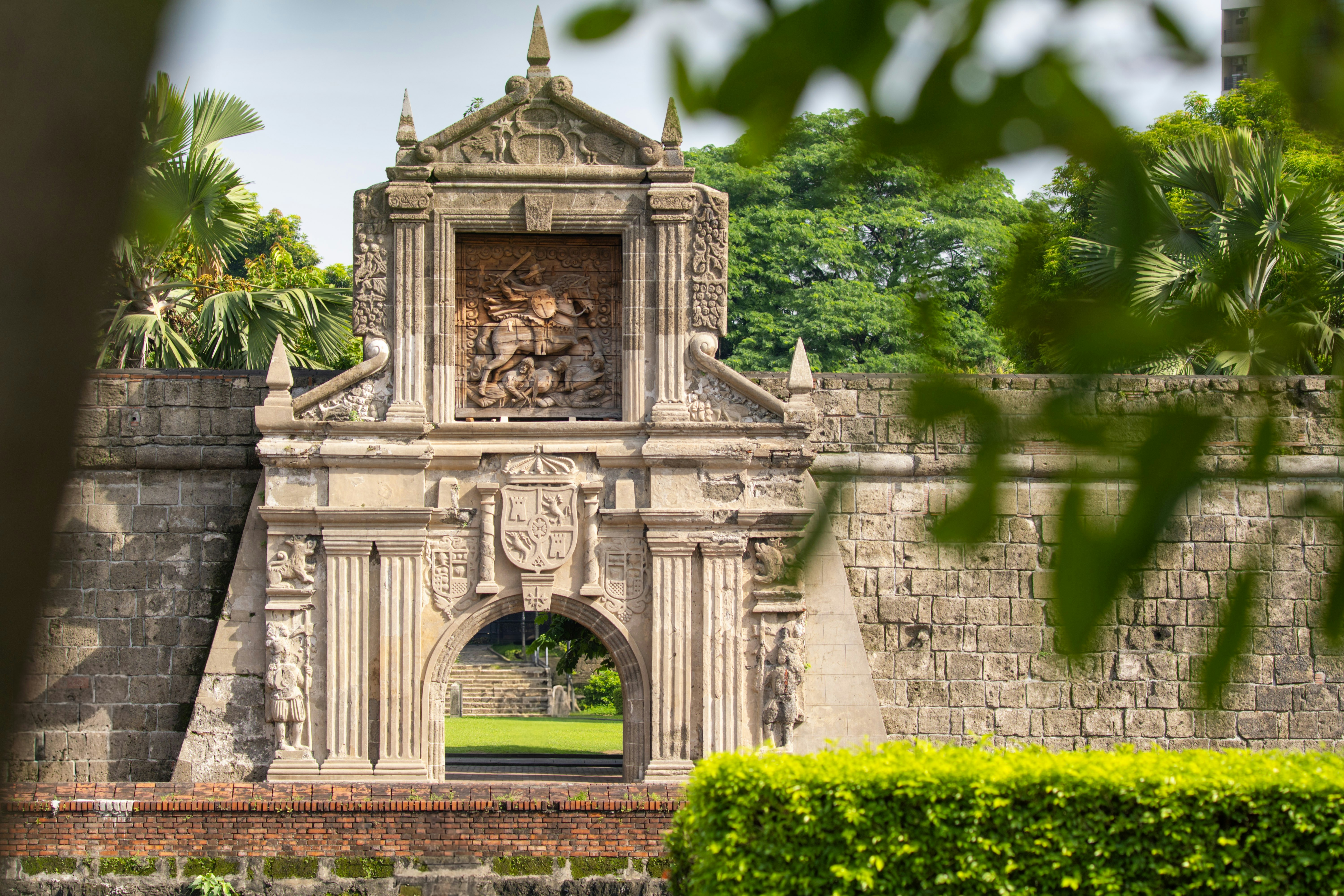 Intricate stone archway adorned with historical reliefs, framed by lush greenery, leading to a serene garden space.