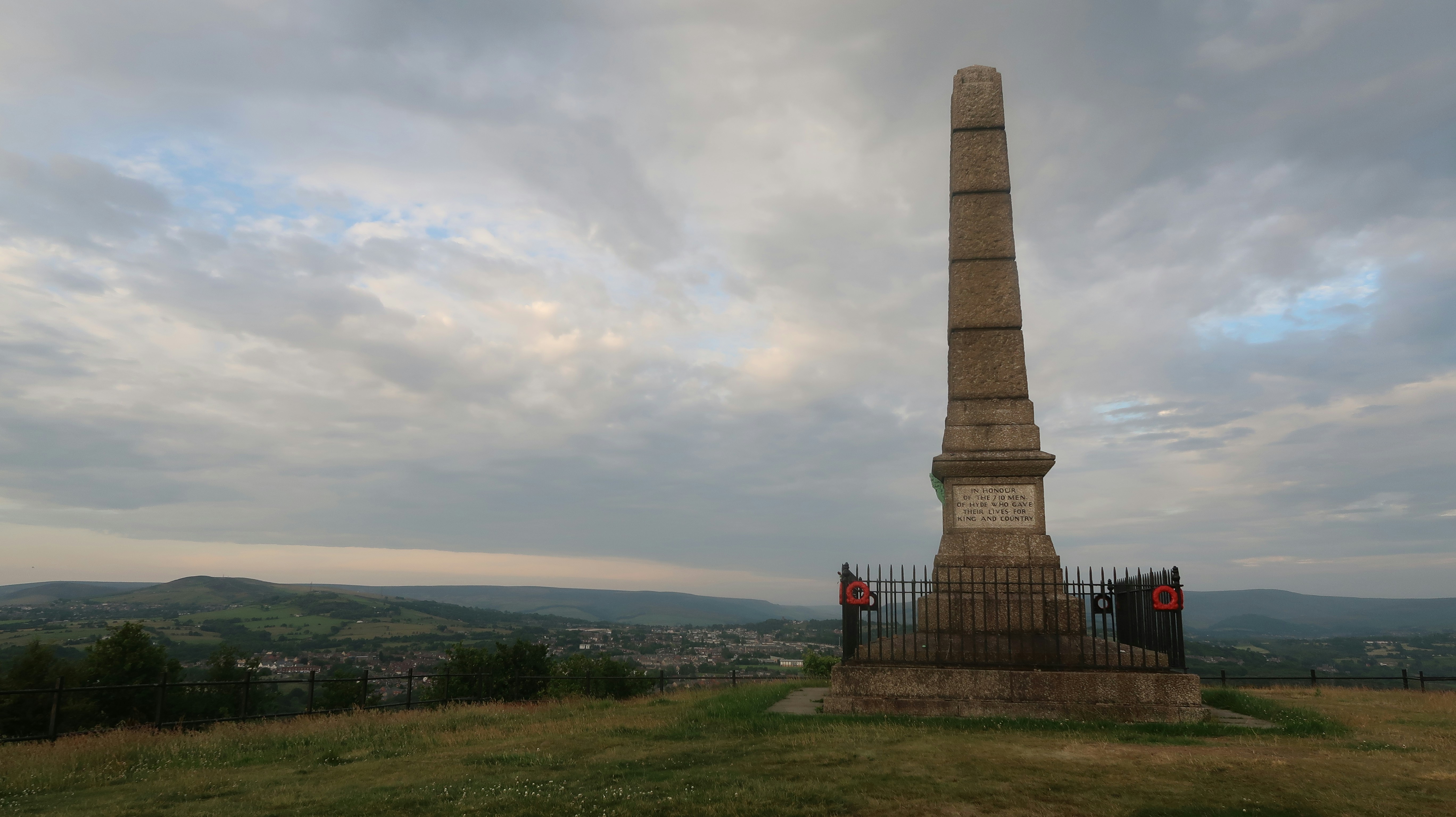 A tall stone monument stands on a hilltop, surrounded by a scenic landscape under a cloudy sky, adorned with memorial wreaths.