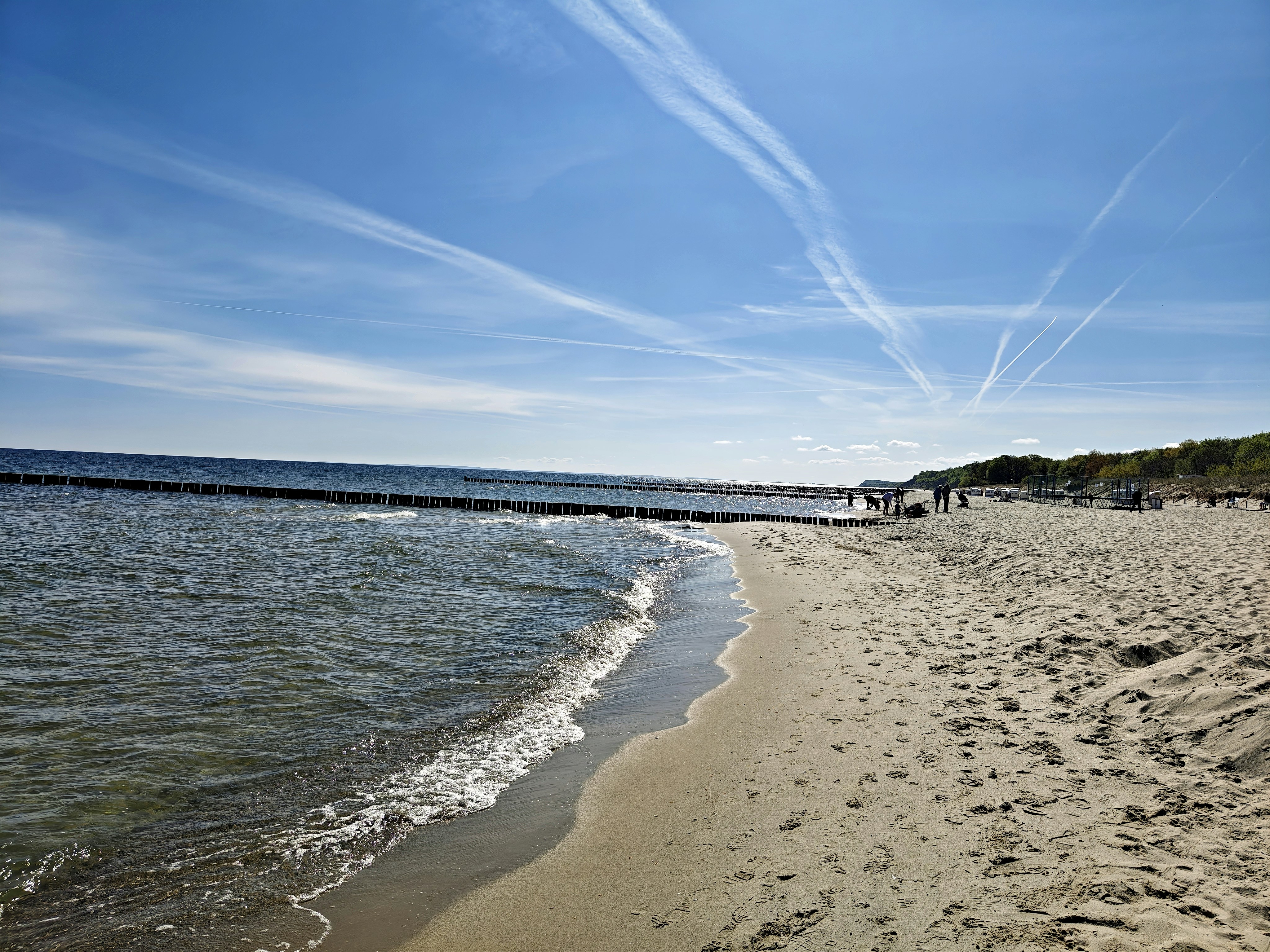 Gentle waves lapping against a sandy beach under a clear blue sky, with a wooden pier extending into the water. People enjoy the serene coastal atmosphere.