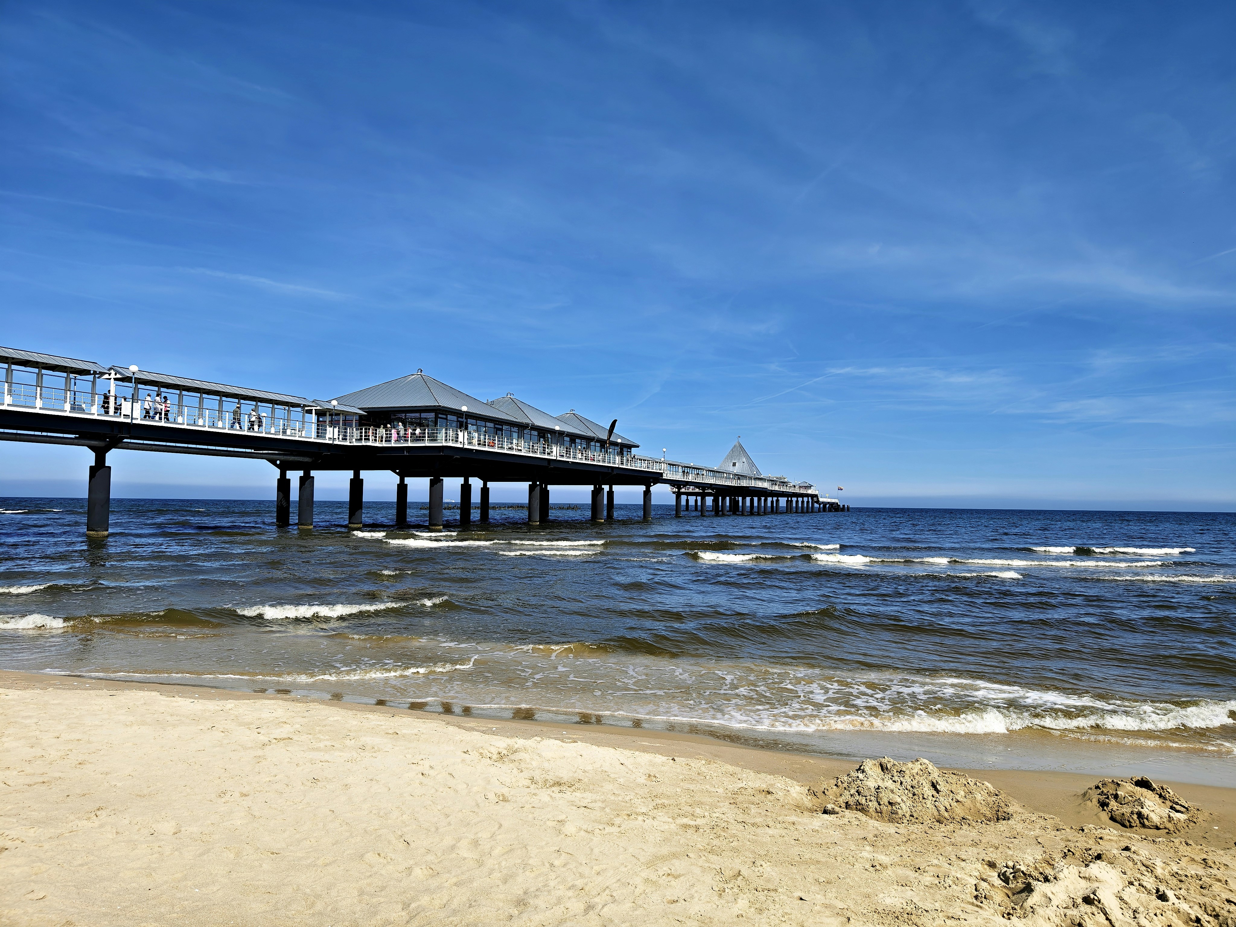 Wooden pier extending over tranquil waters, framed by gentle waves and sandy beach under a clear blue sky.