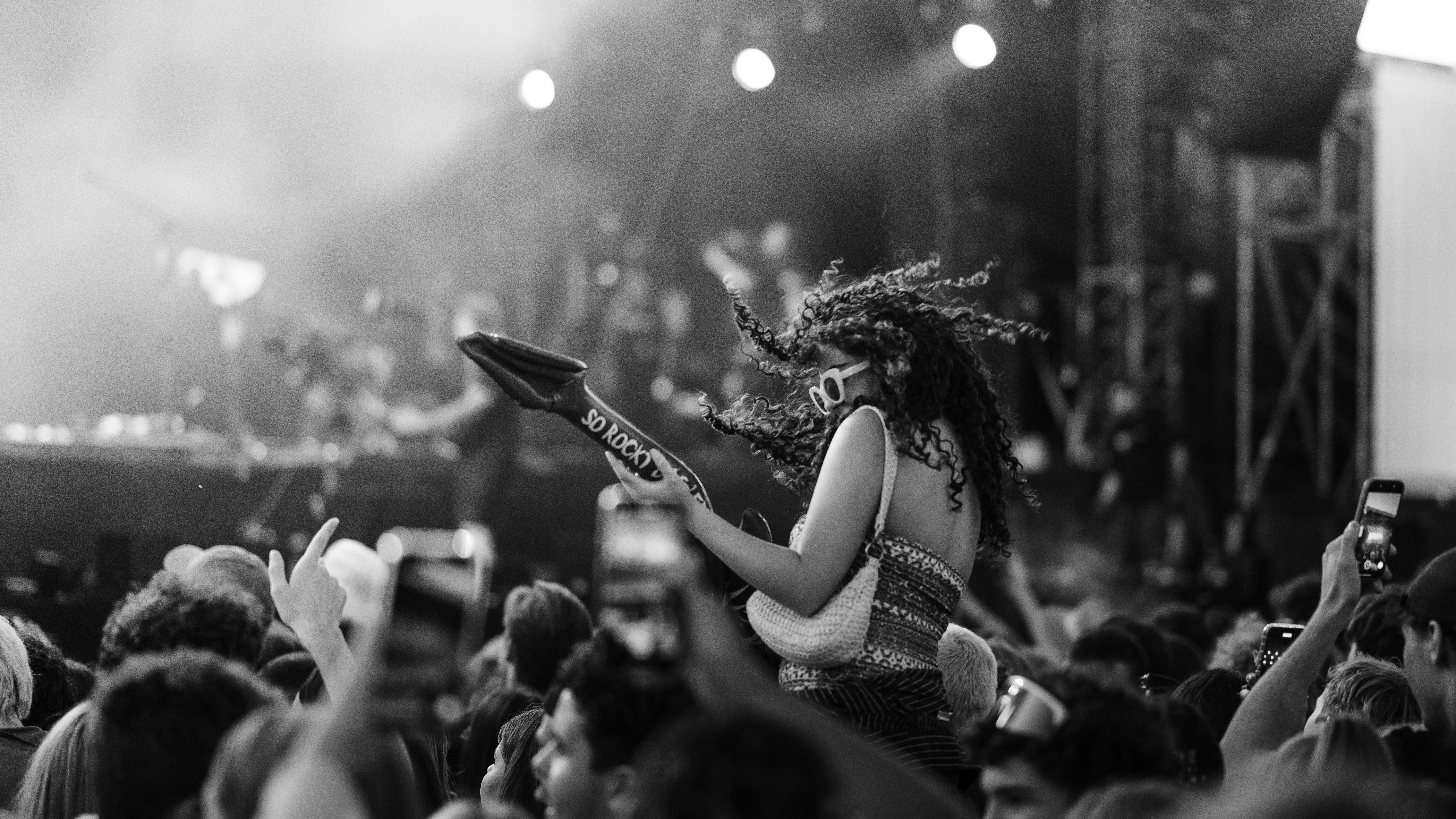 Energetic crowd at a concert with a woman joyfully dancing and waving a large inflatable instrument. The scene is captured in striking black and white.