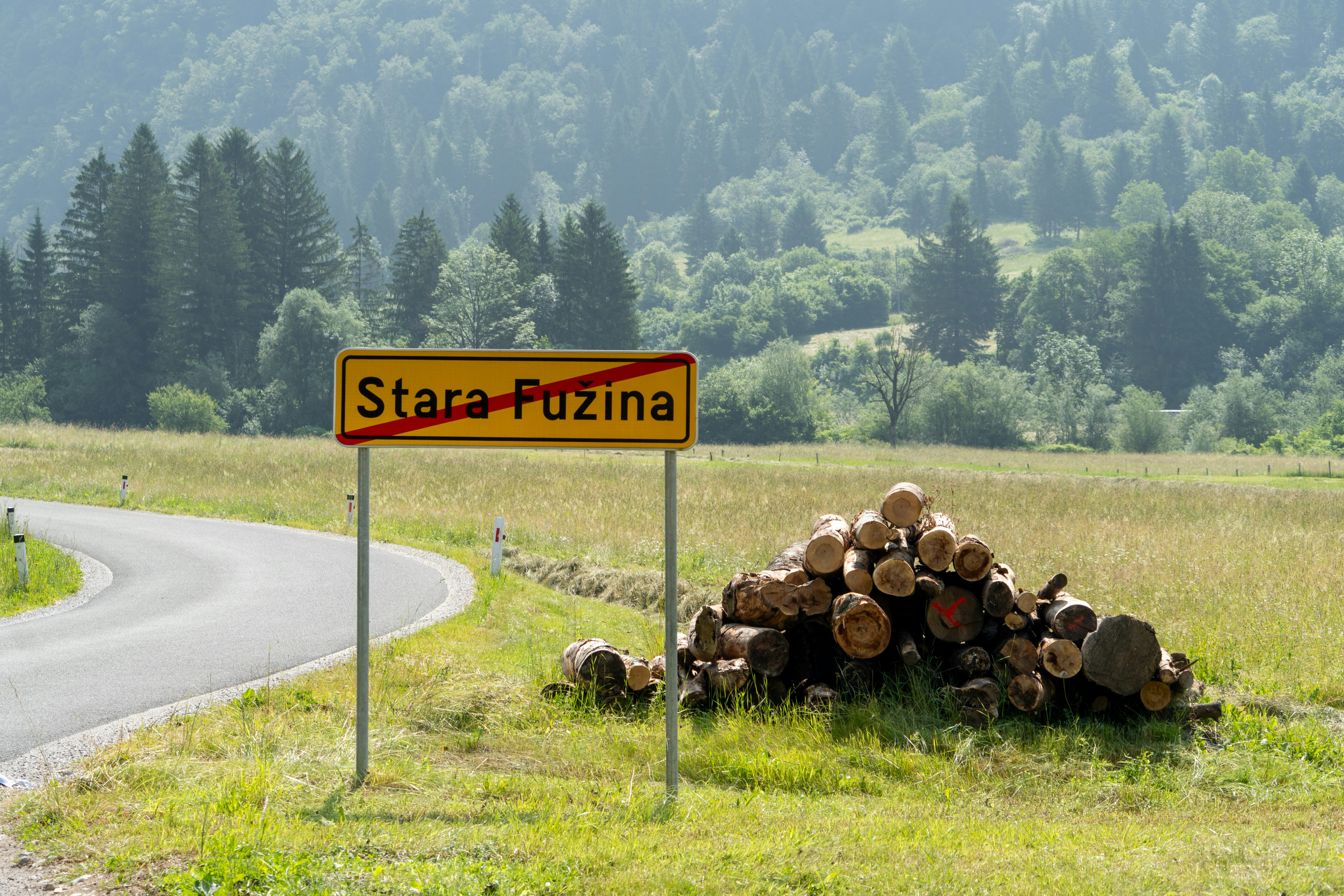 A road sign marks the exit from Stara Fužina, a quiet village in Slovenia, set against a sunlit alpine meadow and forested hills. A neat stack of freshly cut logs rests beside the winding rural road, reflecting the region’s connection to nature and tradition. | A road sign of a town with a stack of logs.