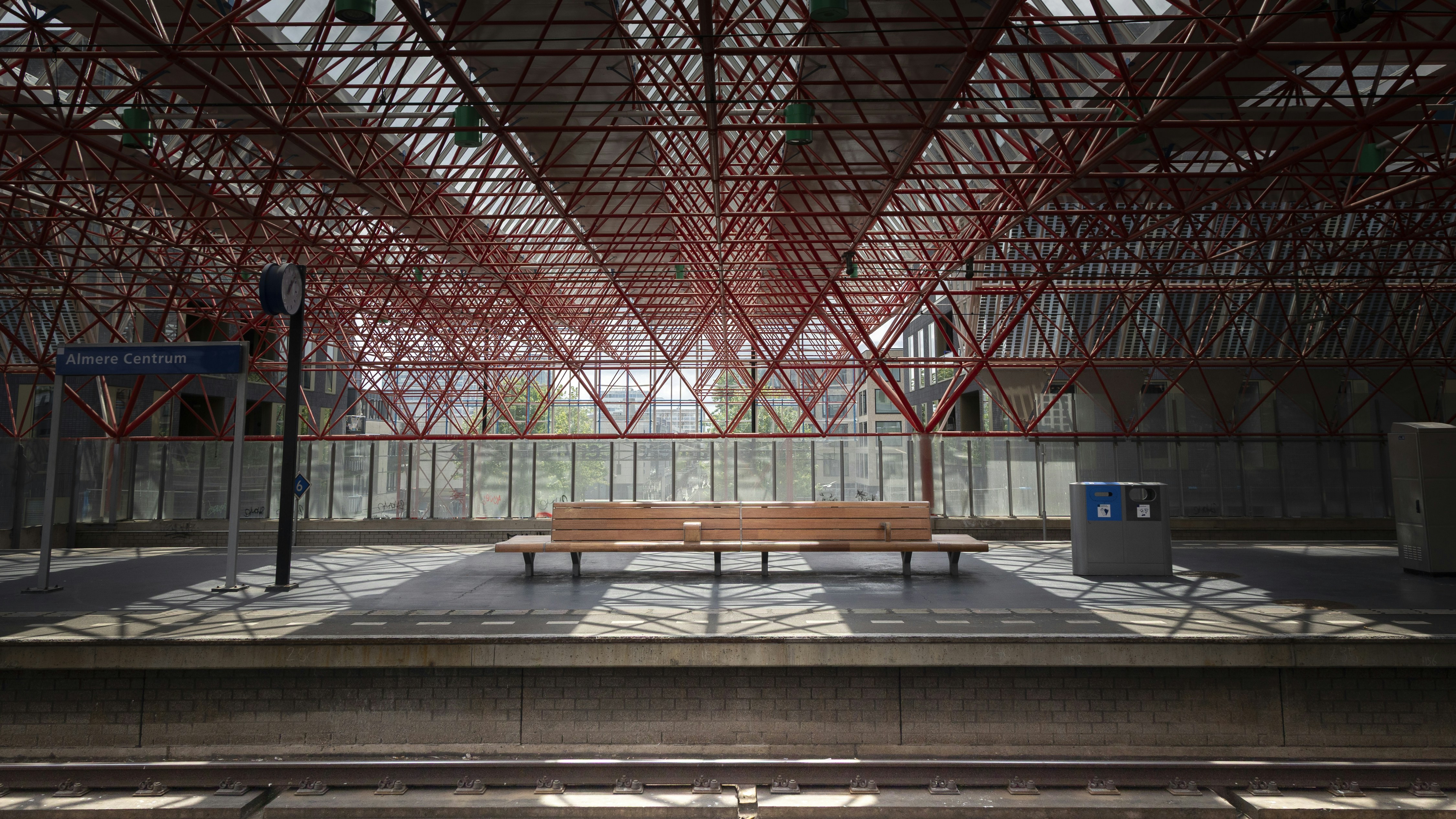 Modern train station interior showcasing an intricate red metal framework and a solitary wooden bench, illuminated by natural light filtering through the glass roof.